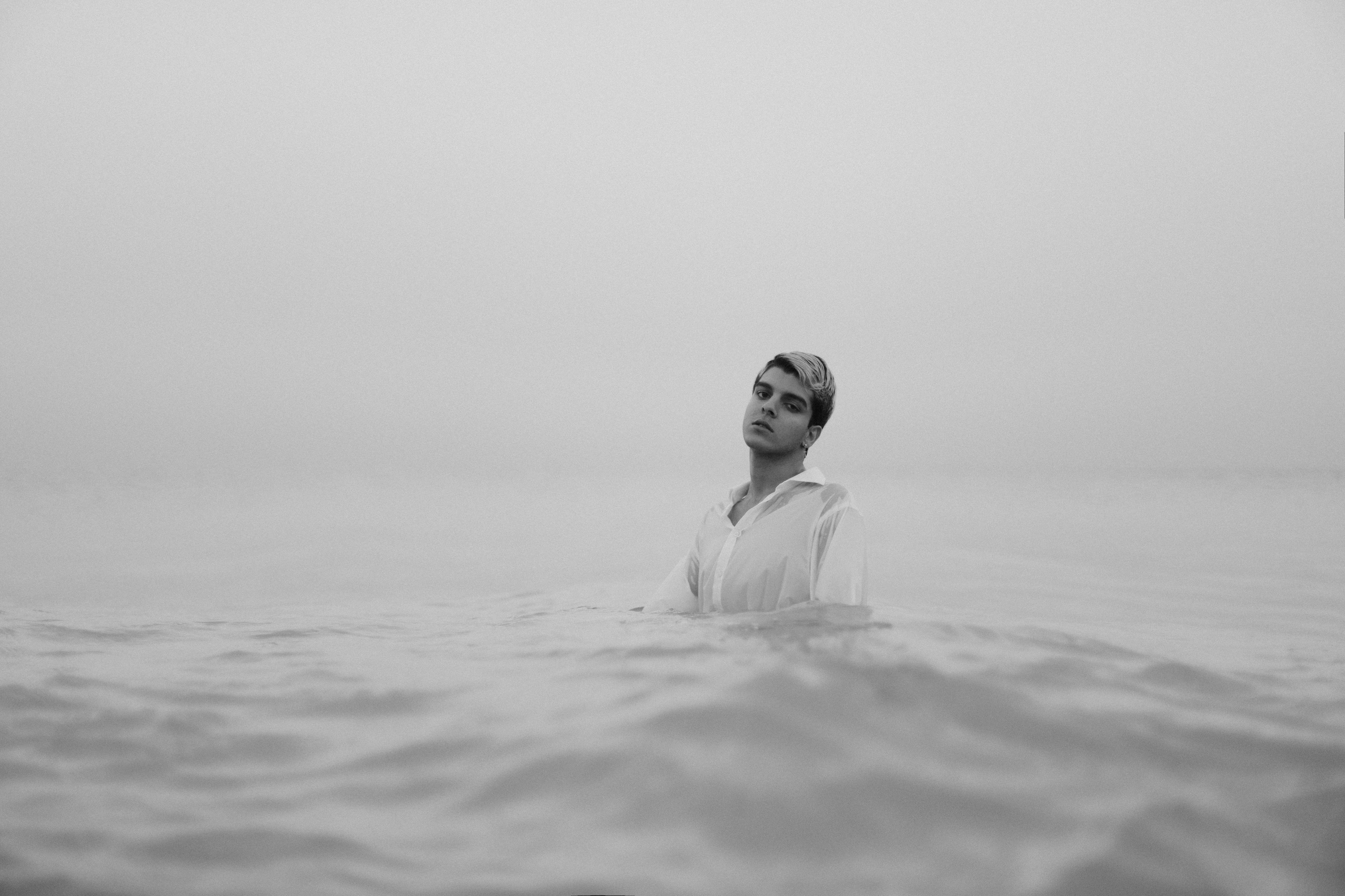 Person standing waist-deep in calm sea, wearing a white shirt, with a misty horizon in the background.