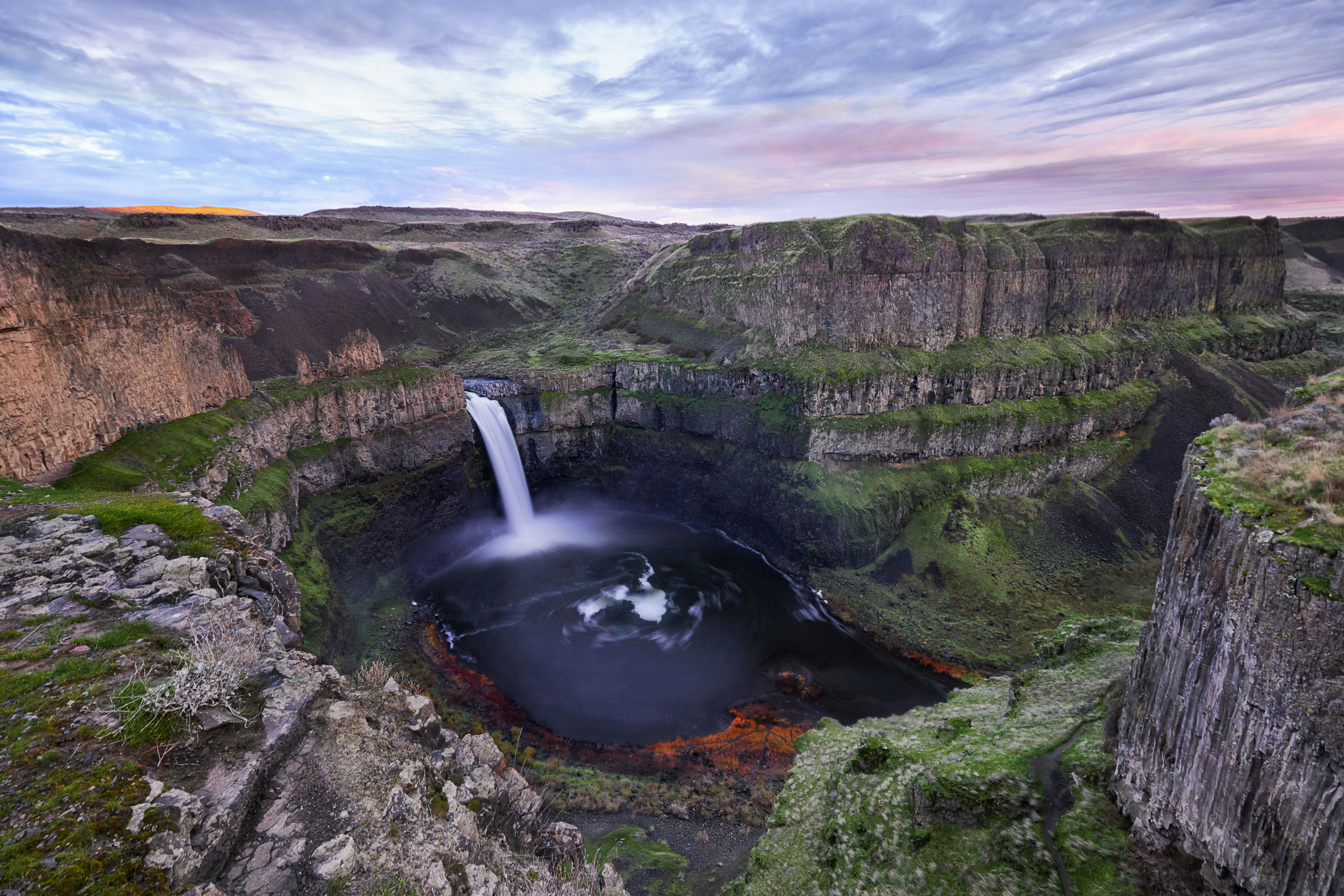 Waterfall cascading into a circular pool surrounded by rugged cliffs and lush greenery under a pastel sky.