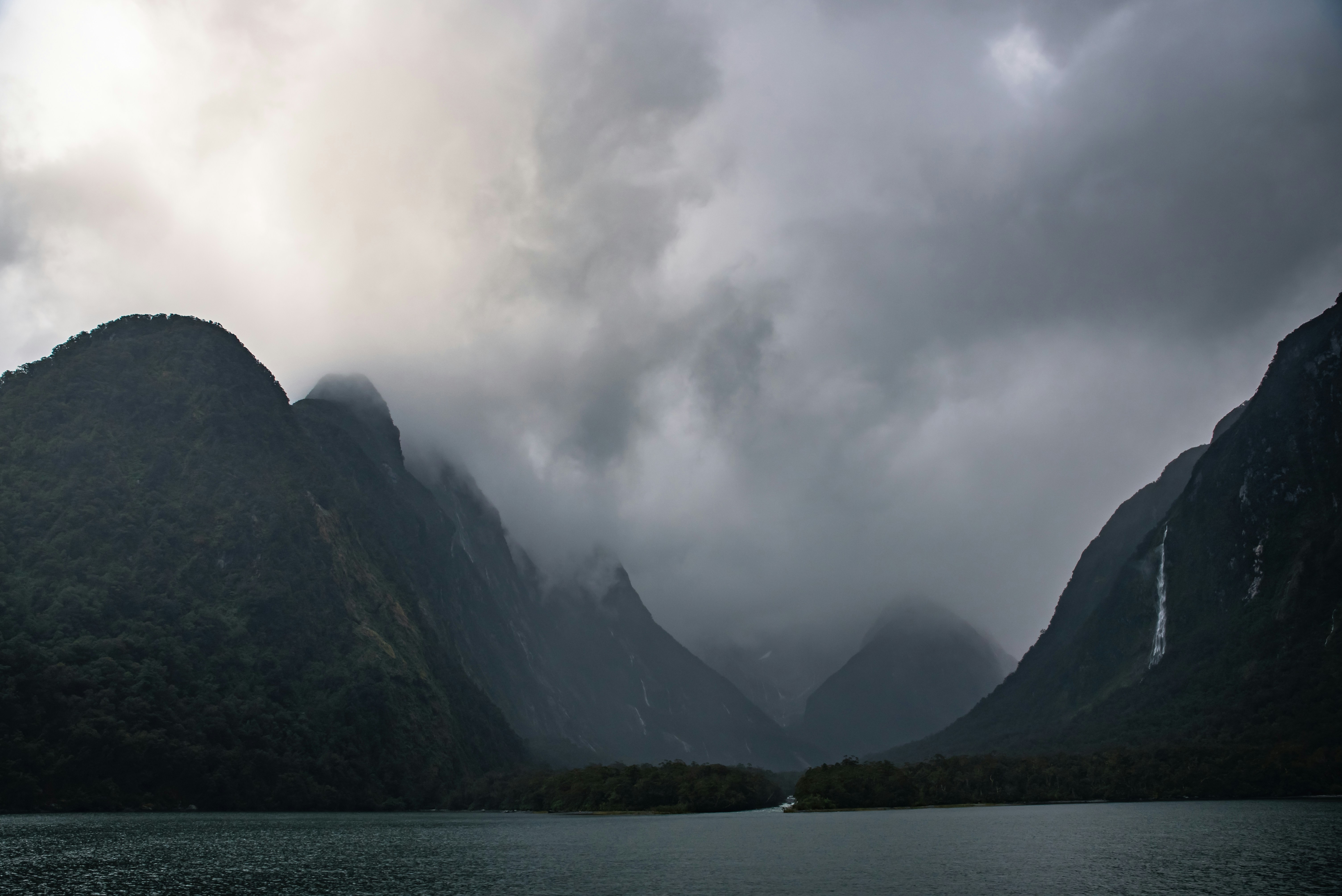 Dramatic cloud-covered mountains towering over a tranquil lake with a distant waterfall.