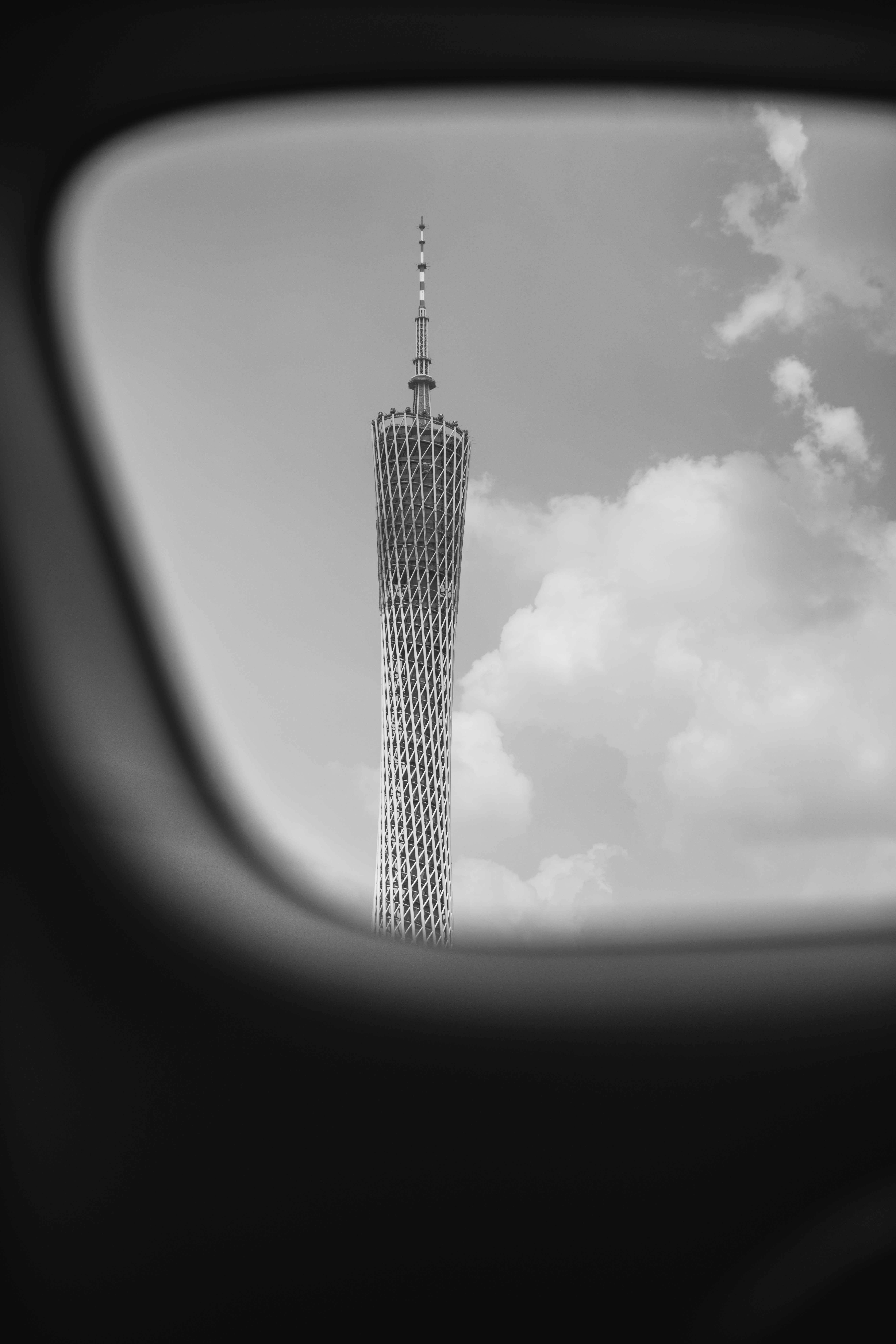 A striking view of a tall tower framed by a vehicle's window, with a backdrop of fluffy clouds. The monochrome tones enhance the architectural details.
