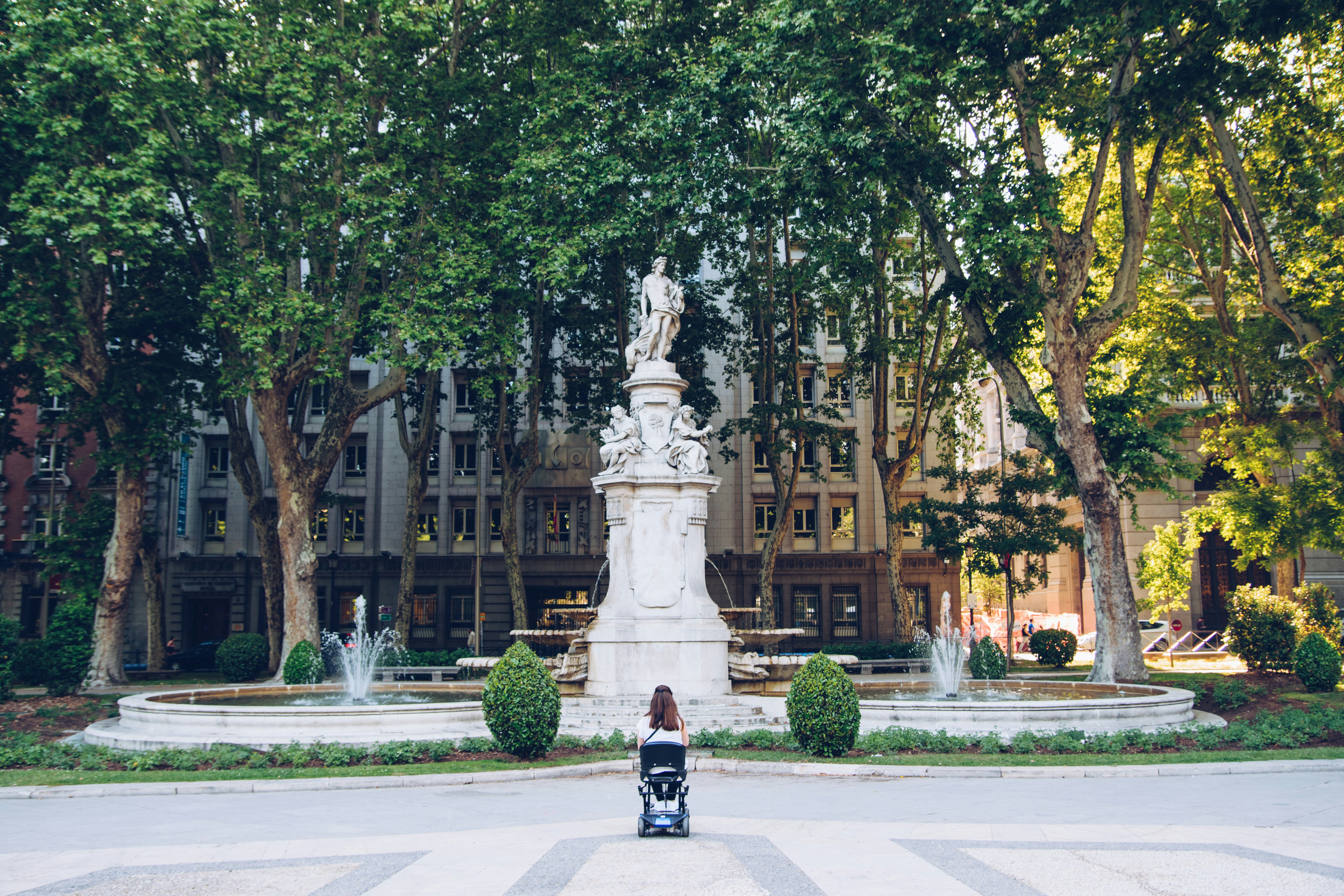 A tranquil scene featuring a person seated in front of a grand fountain and statue, framed by lush greenery in an urban park.