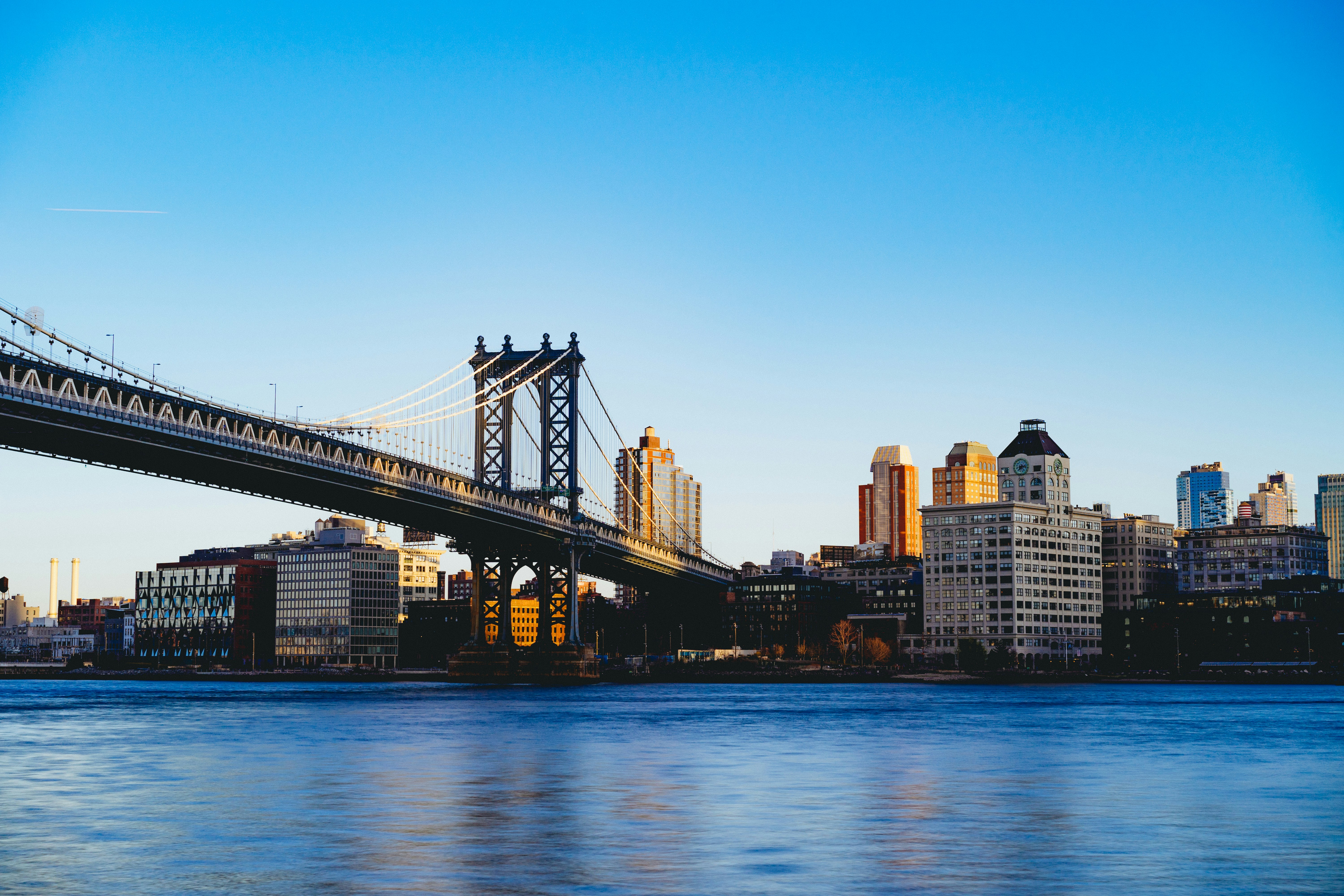 Suspension bridge spanning a river with a city skyline under a clear blue sky.