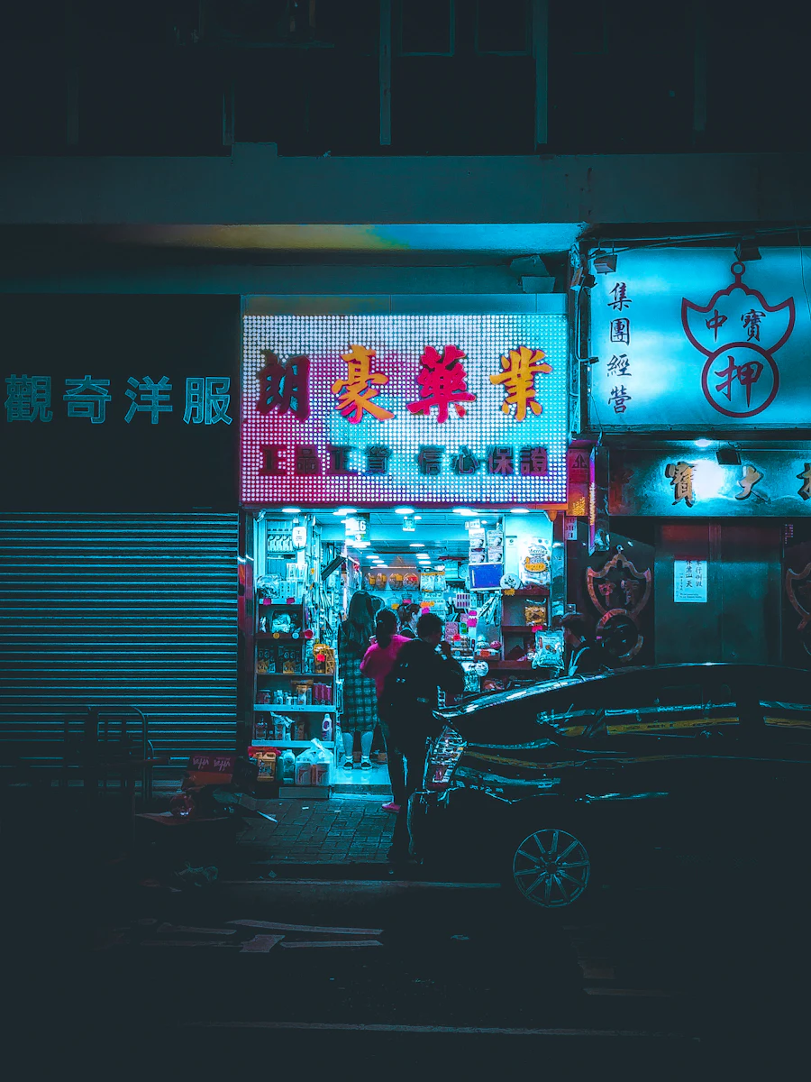 A small, brightly lit shop with Chinese signage glows against a dark street at night, with a few people standing at the entrance. A parked car fills the lower right foreground.