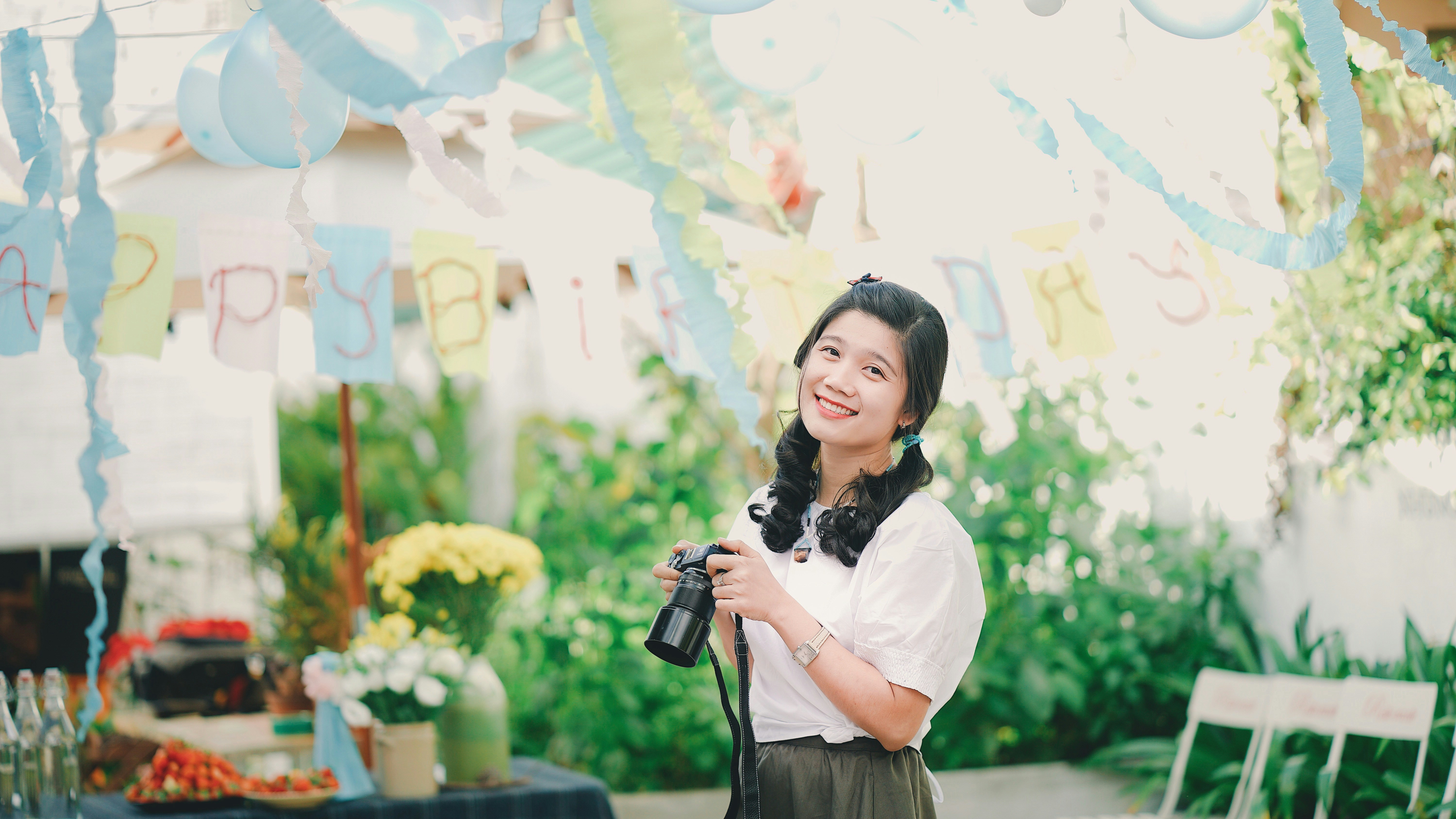 Woman smiling with a camera amidst colorful party decorations in a garden.