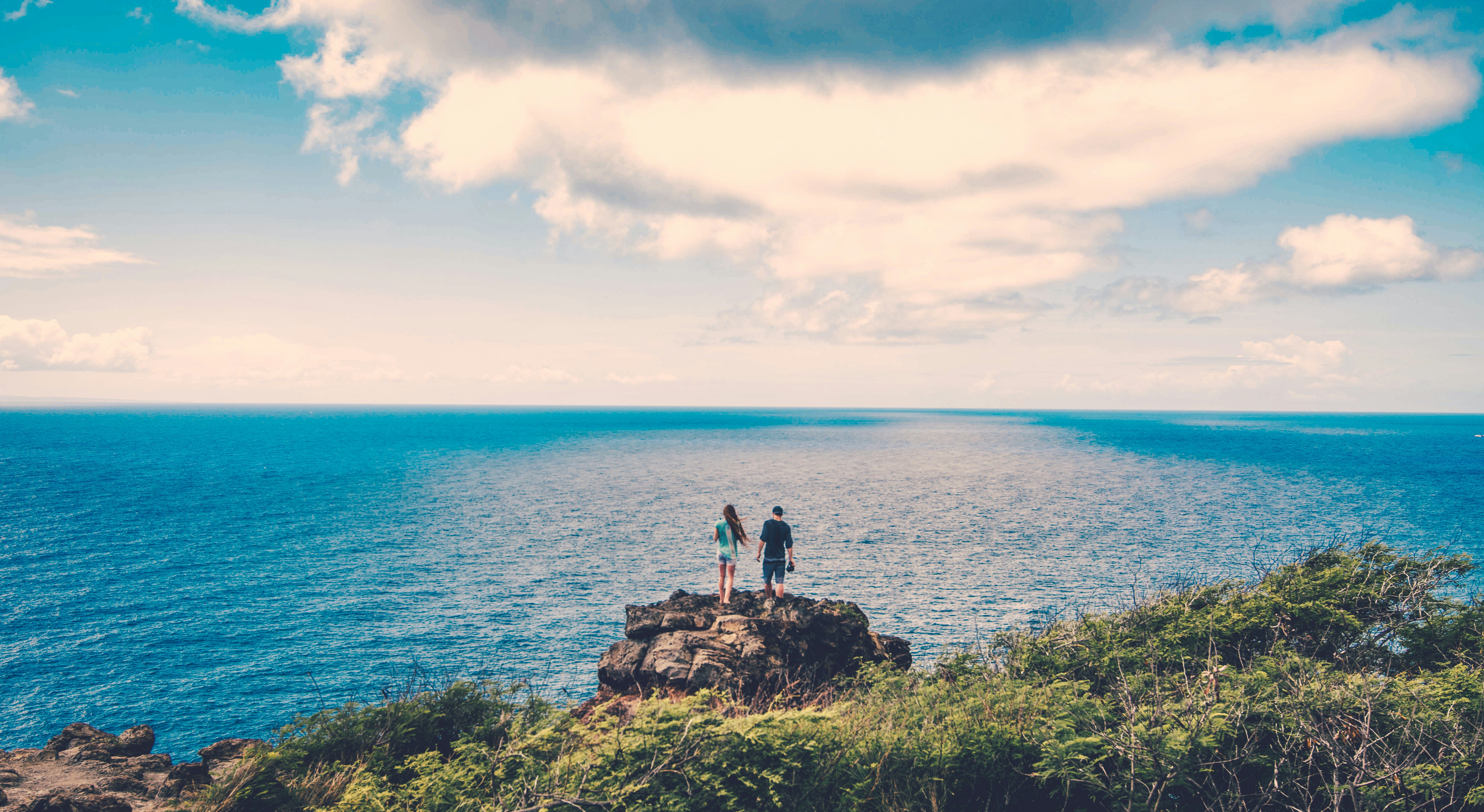 Two people standing on a cliff overlooking a vast ocean under a partly cloudy sky.