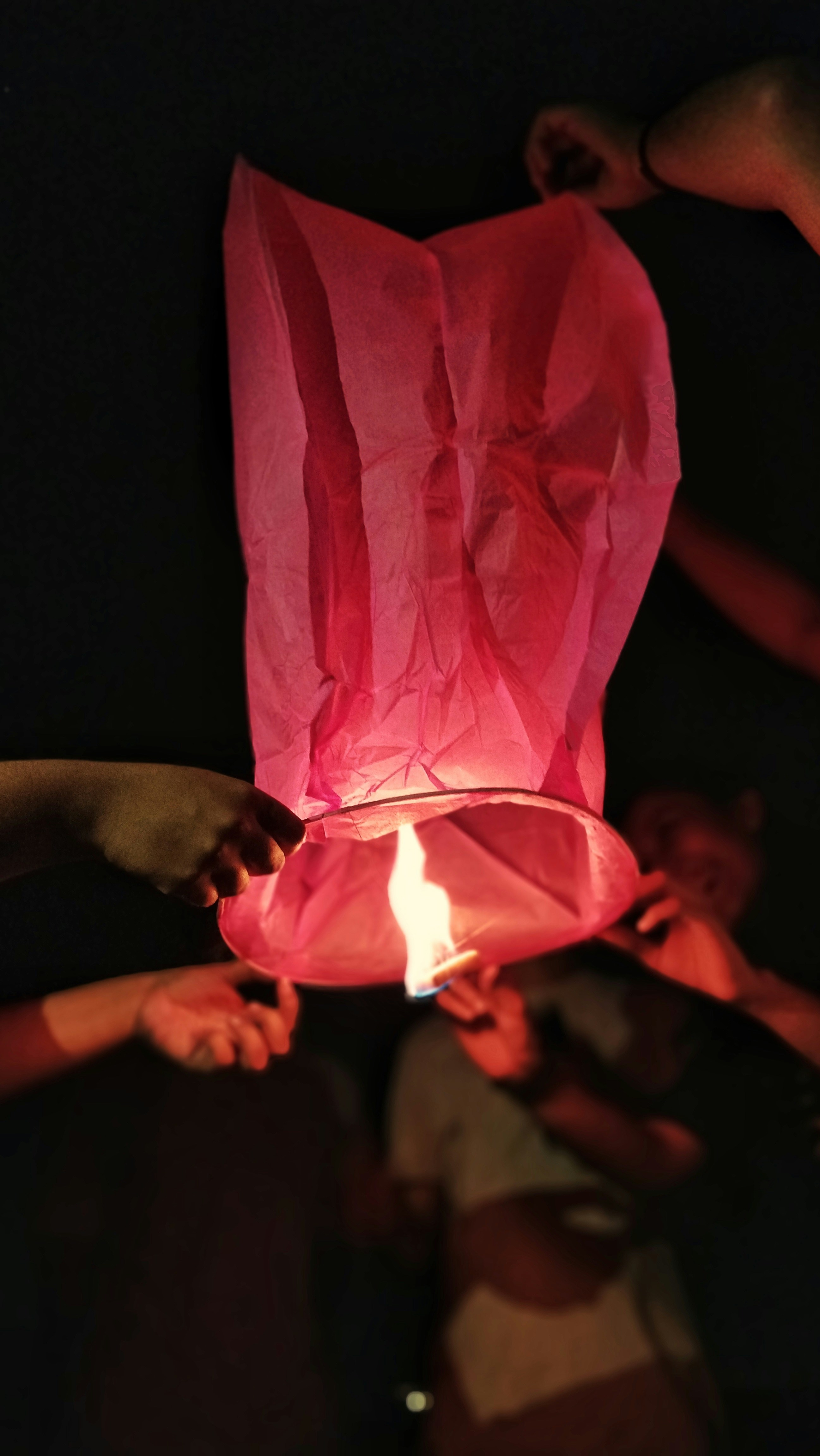 A pink paper lantern glows as a flame lights its crinkled folds, held by several hands against a dark backdrop.