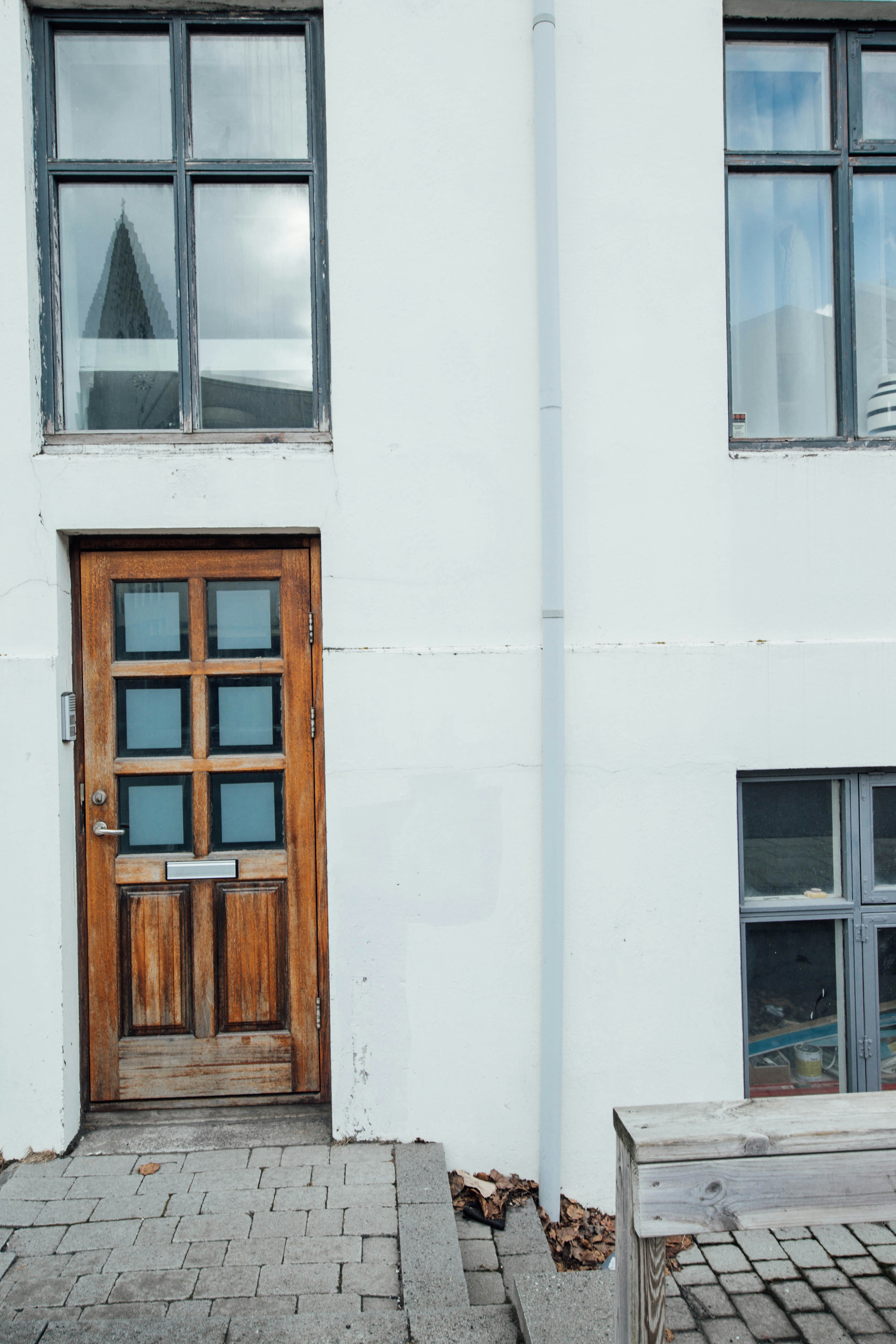A wooden door with glass panels stands against a white wall, framed by large windows reflecting the sky. The scene captures a blend of architecture and simplicity.