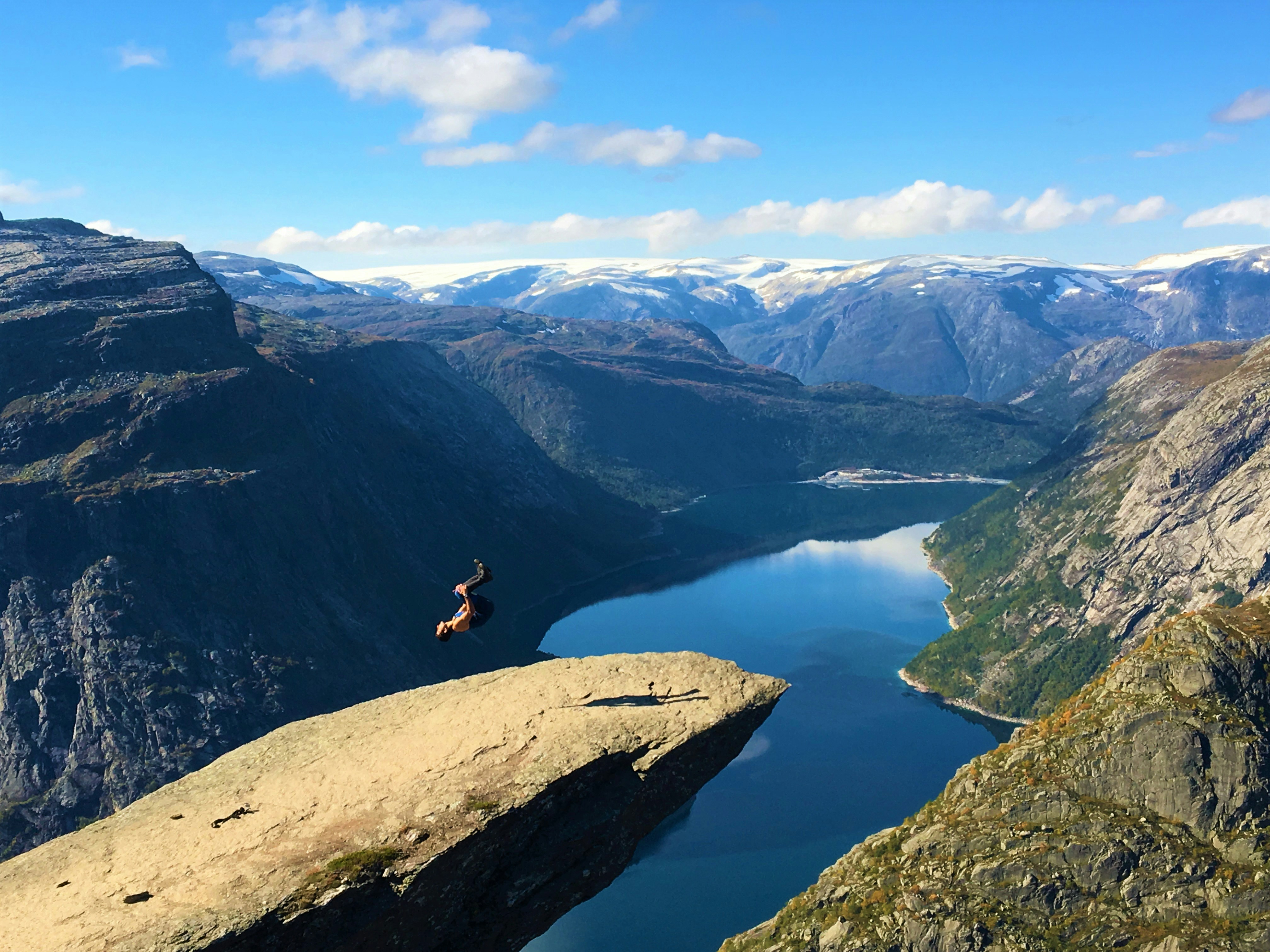 Person mid-jump on the edge of Trolltunga with expansive mountain and fjord view.