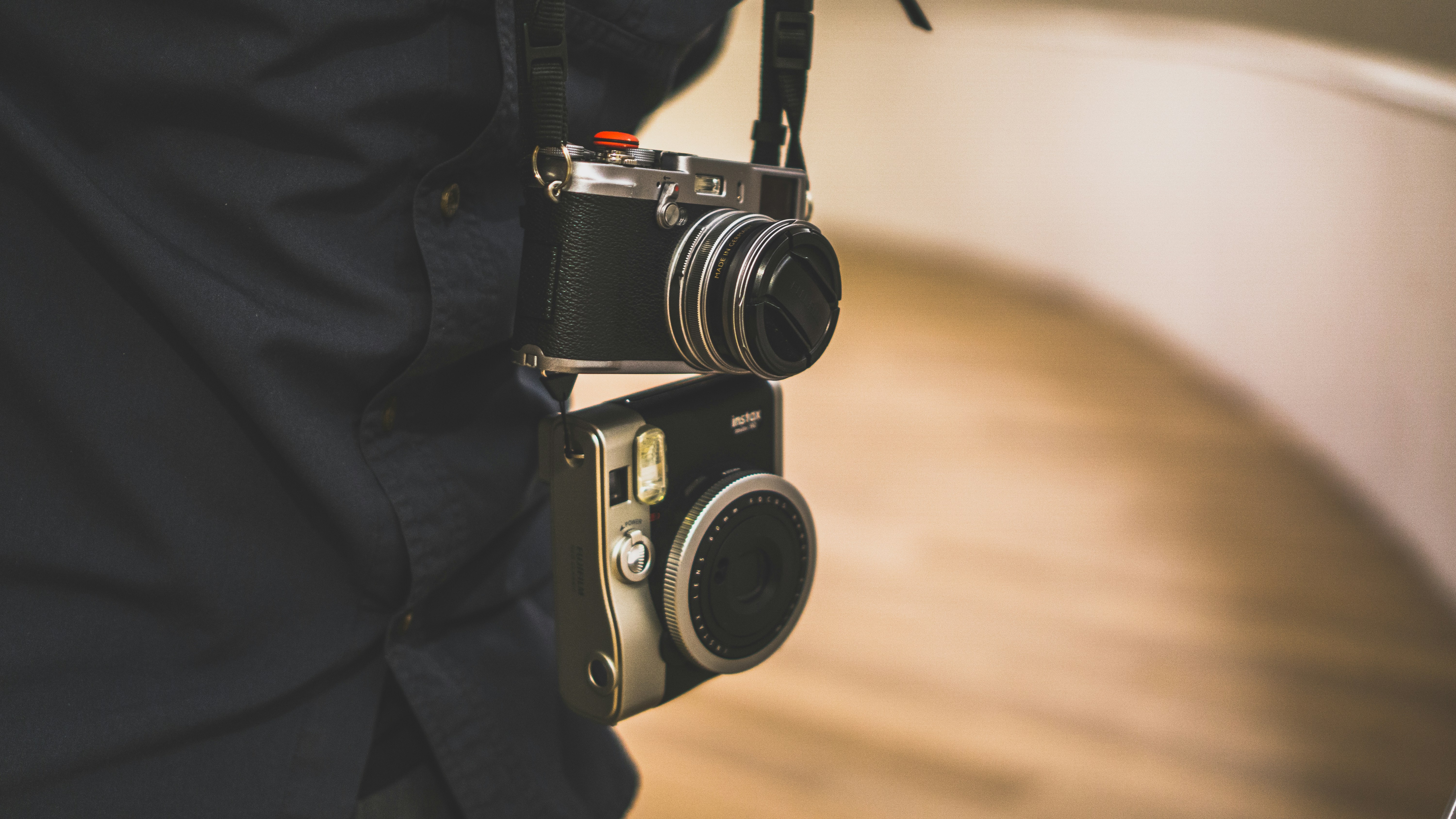 Two cameras hanging from a person's waist, showcasing a blend of modern and vintage design elements. The setting features a softly blurred background that emphasizes the equipment.