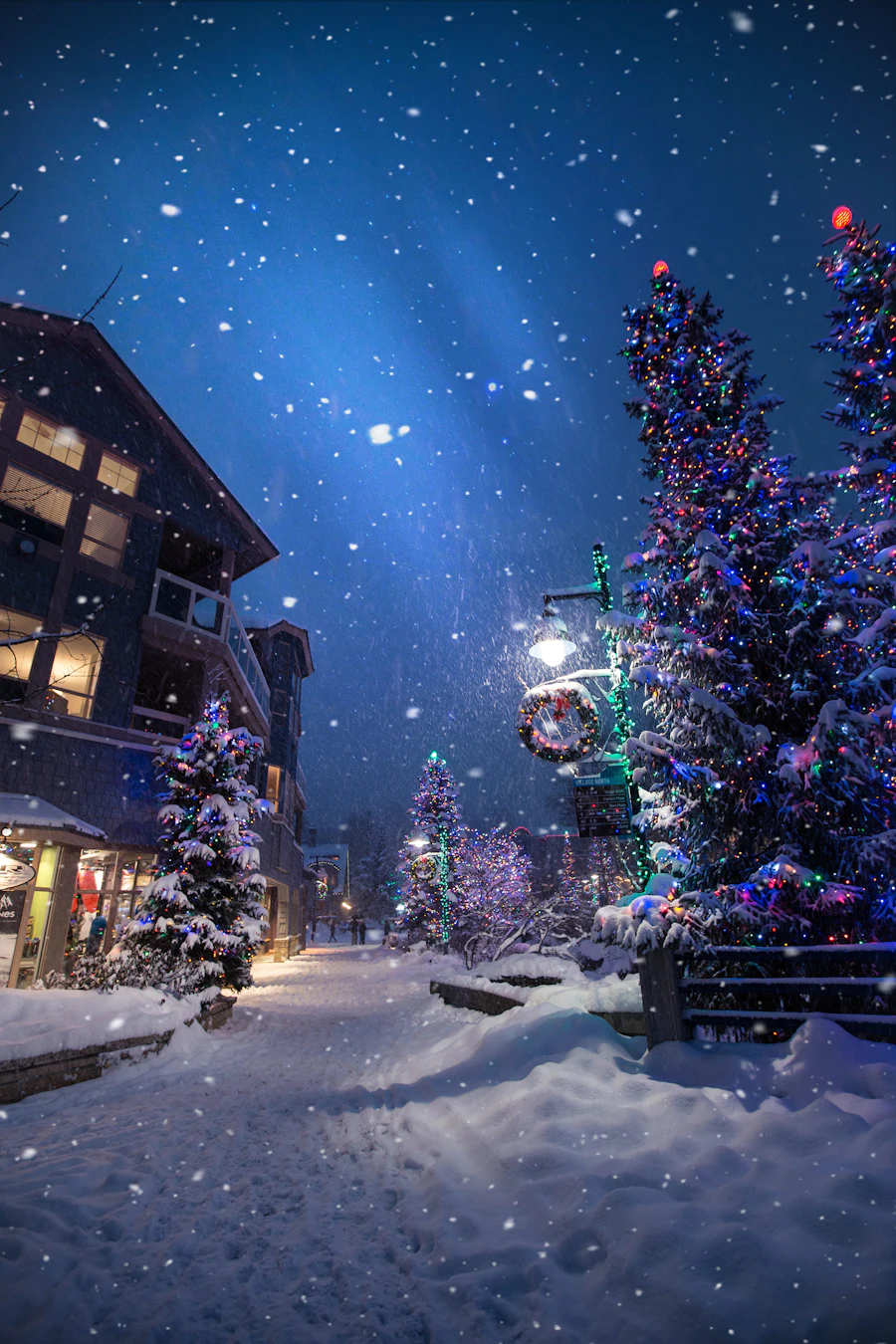 Snow falls over a narrow village street lined with decorated Christmas trees and glowing shopfronts. A deep blue evening sky and warm lights create a festive winter scene.