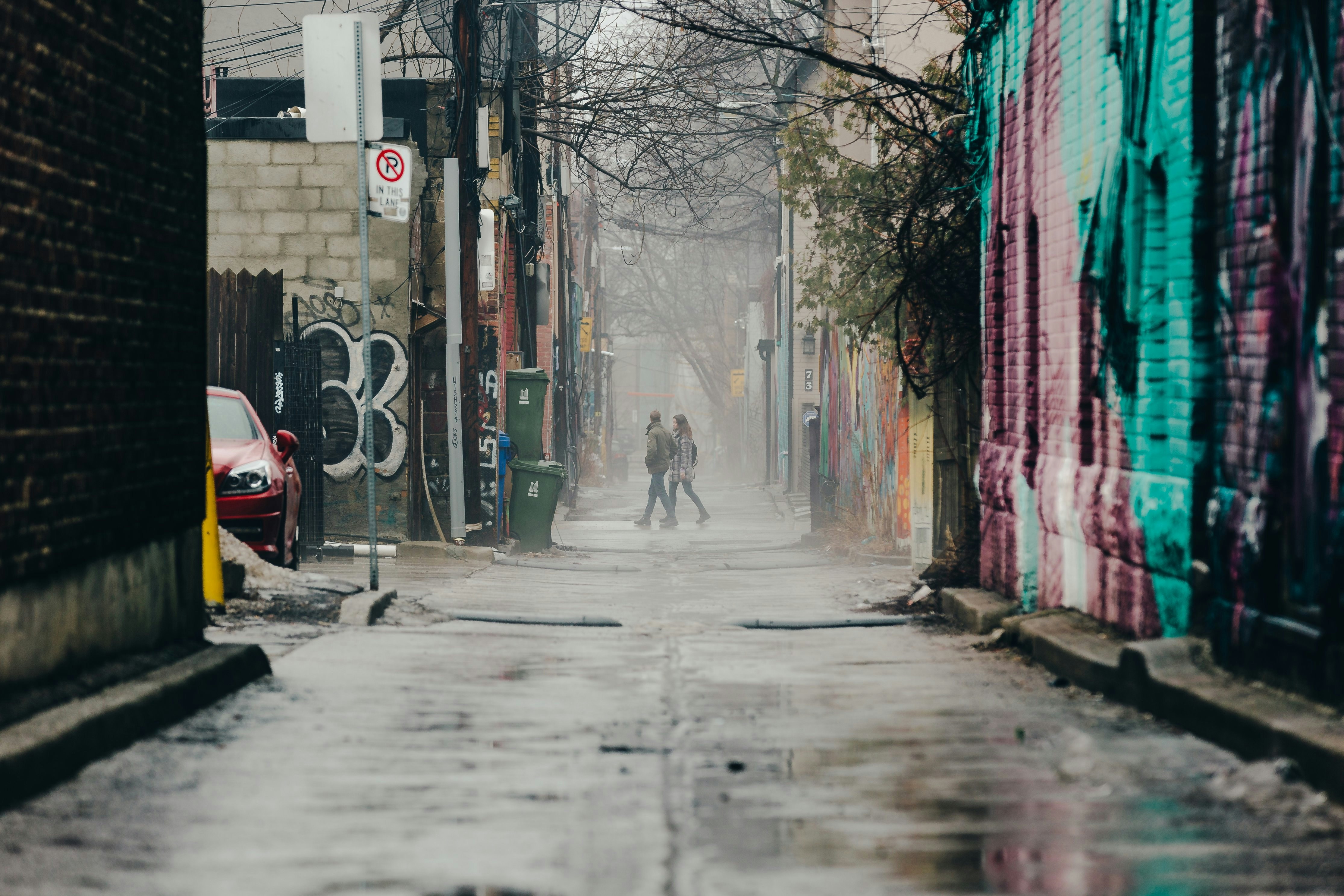 Narrow alley with wet pavement and graffiti-covered walls, two people walking in the distance through morning fog.