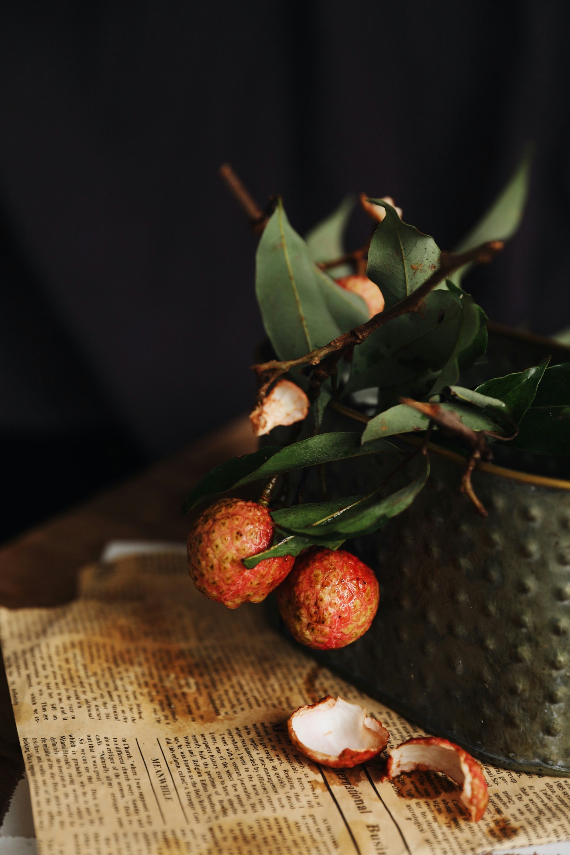 Lychee fruits nestled among vibrant green leaves in a rustic bowl, resting on an old newspaper. The scene evokes a sense of natural abundance.