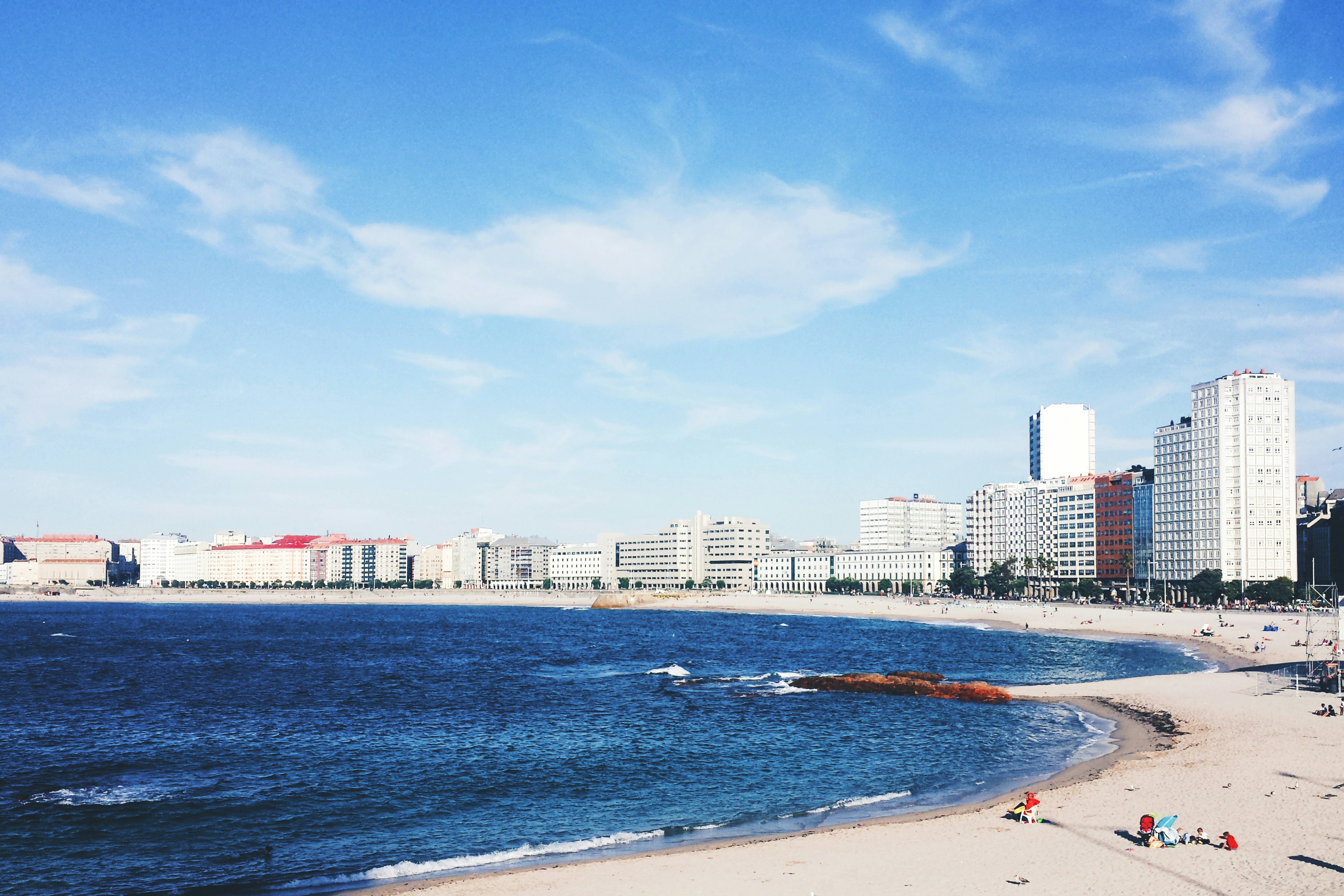 Coastal cityscape with tall white buildings along a sandy beach under a bright blue sky.