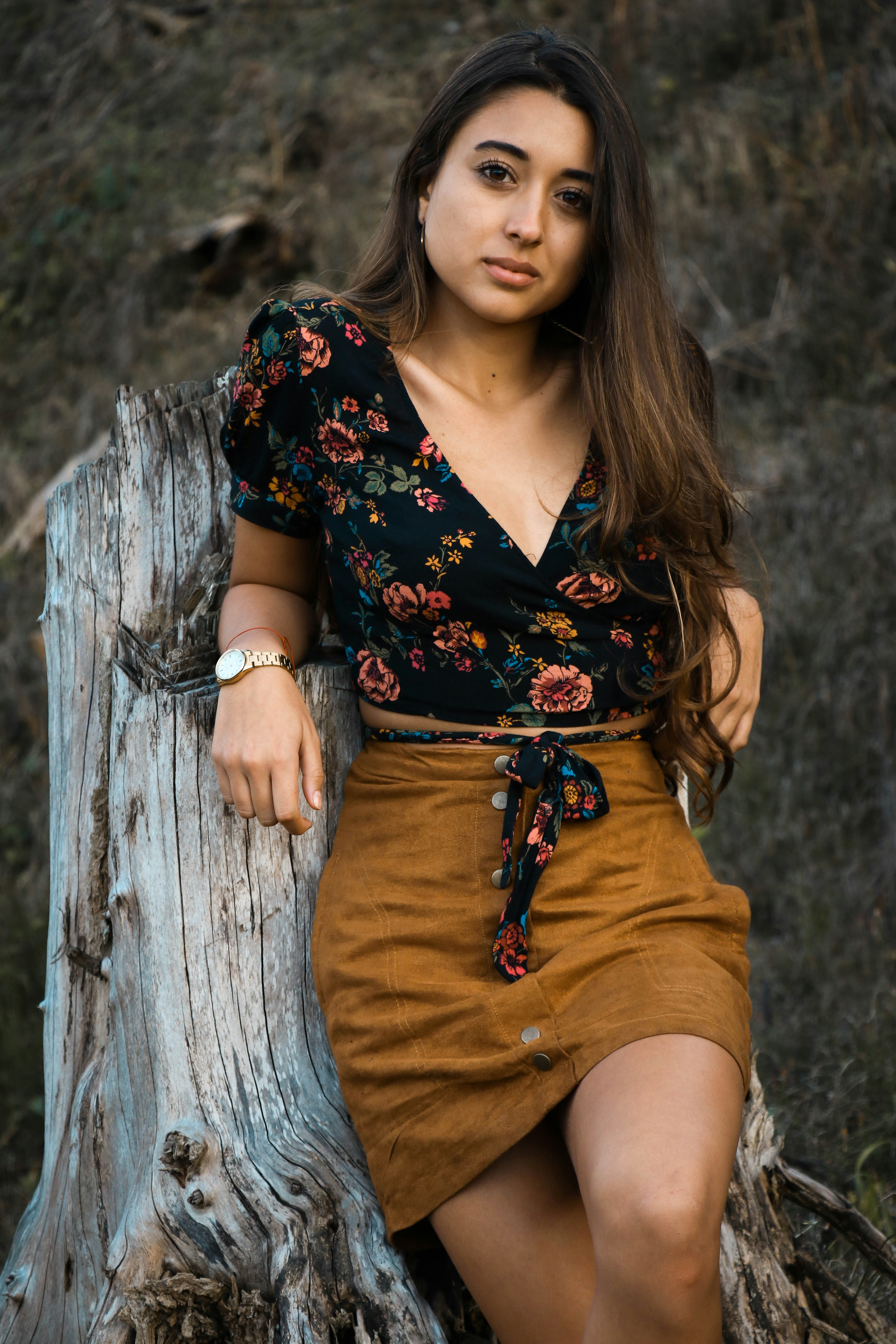 Young woman seated on a weathered tree stump, dressed in a floral top and a tan skirt, exuding a calm and contemplative aura.