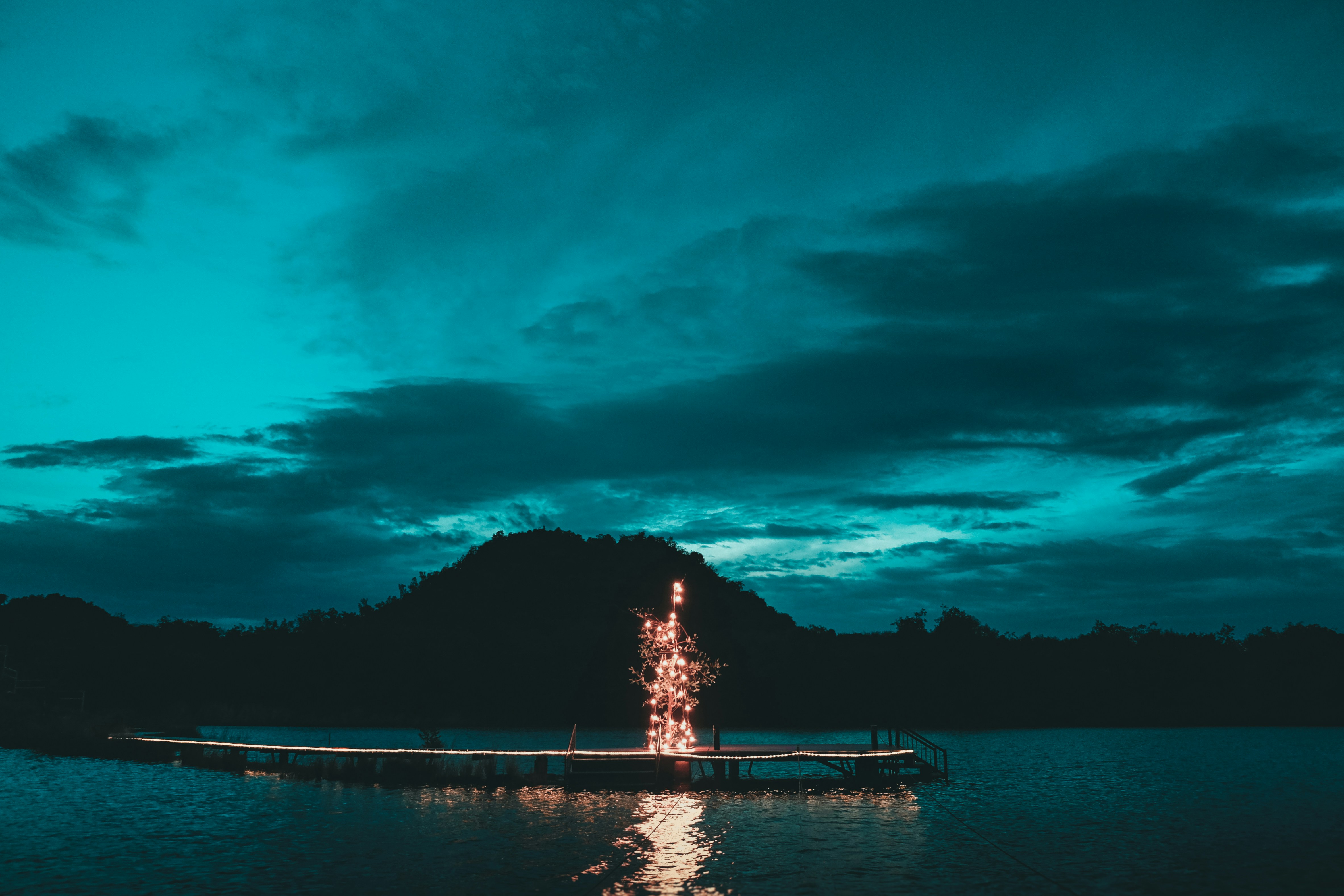 A tranquil scene featuring a glowing structure on a lake, surrounded by dark silhouettes of hills under a moody sky. The interplay of light and shadow creates a serene atmosphere.