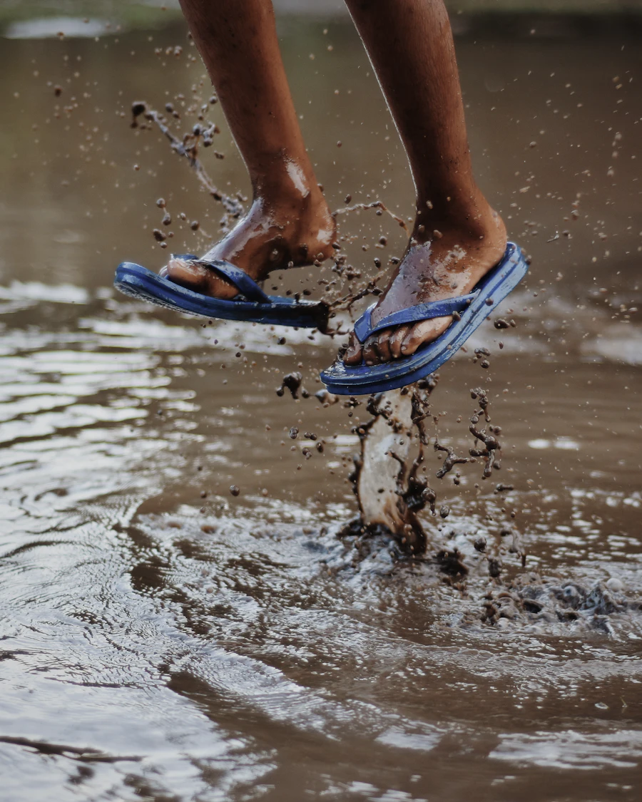 Feet in blue flip-flops leap through muddy water, creating splashes and ripples. The scene captures a playful moment in a natural setting.