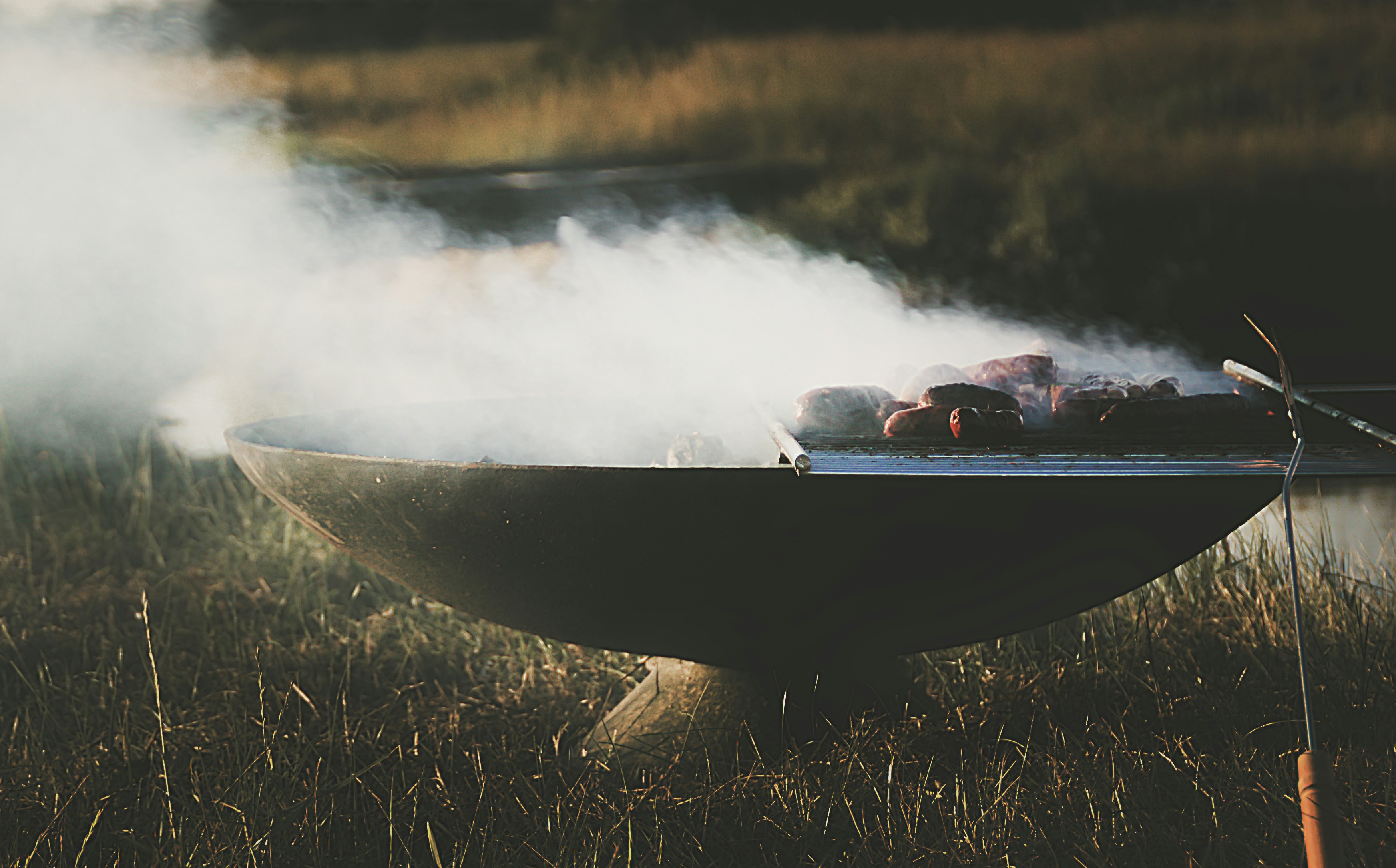 A charcoal grill with sizzling meats surrounded by wisps of smoke, set against a backdrop of tall grass in a serene outdoor setting.