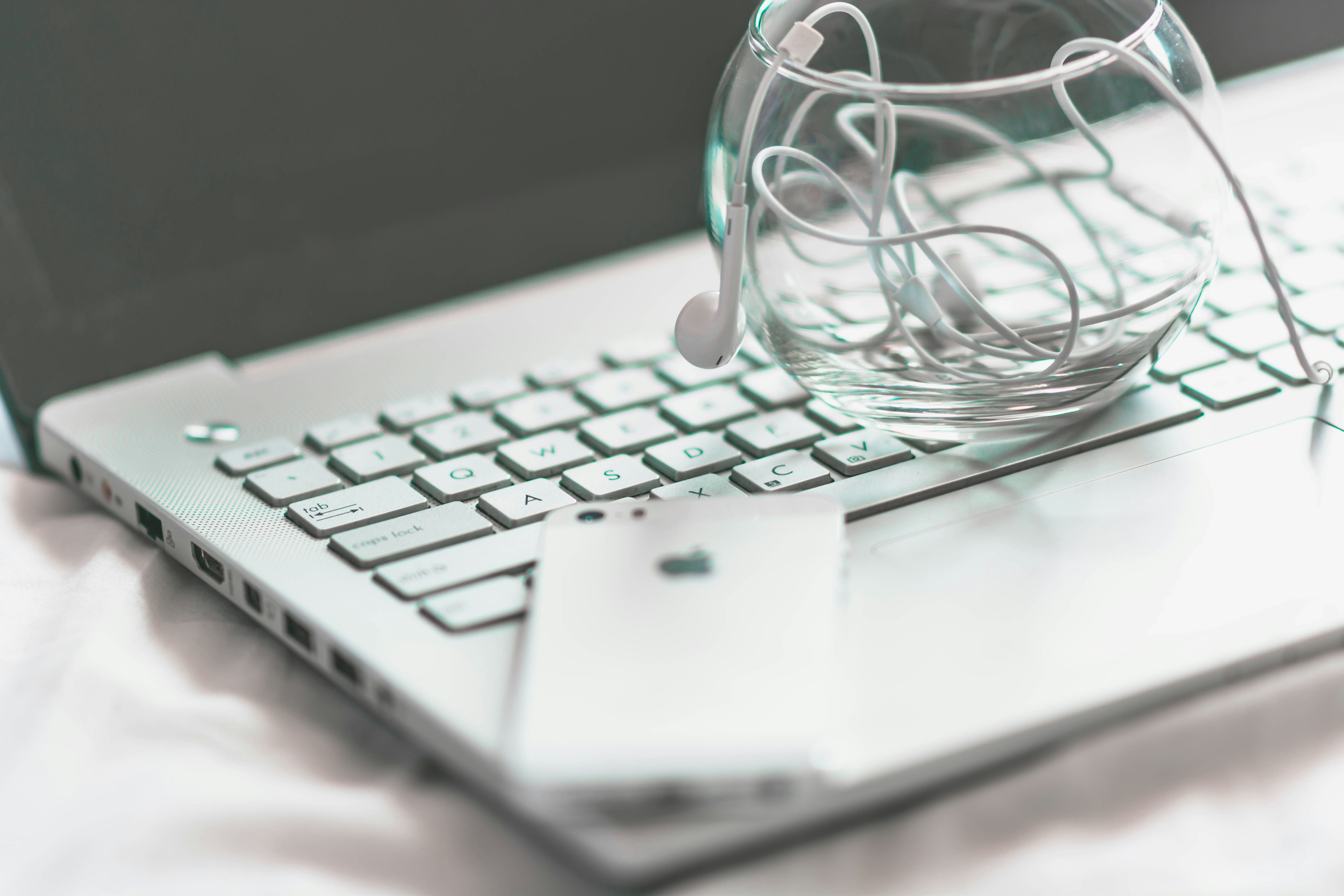A sleek laptop keyboard with a glass bowl containing tangled earphones and a smartphone nearby, showcasing a modern workspace aesthetic.