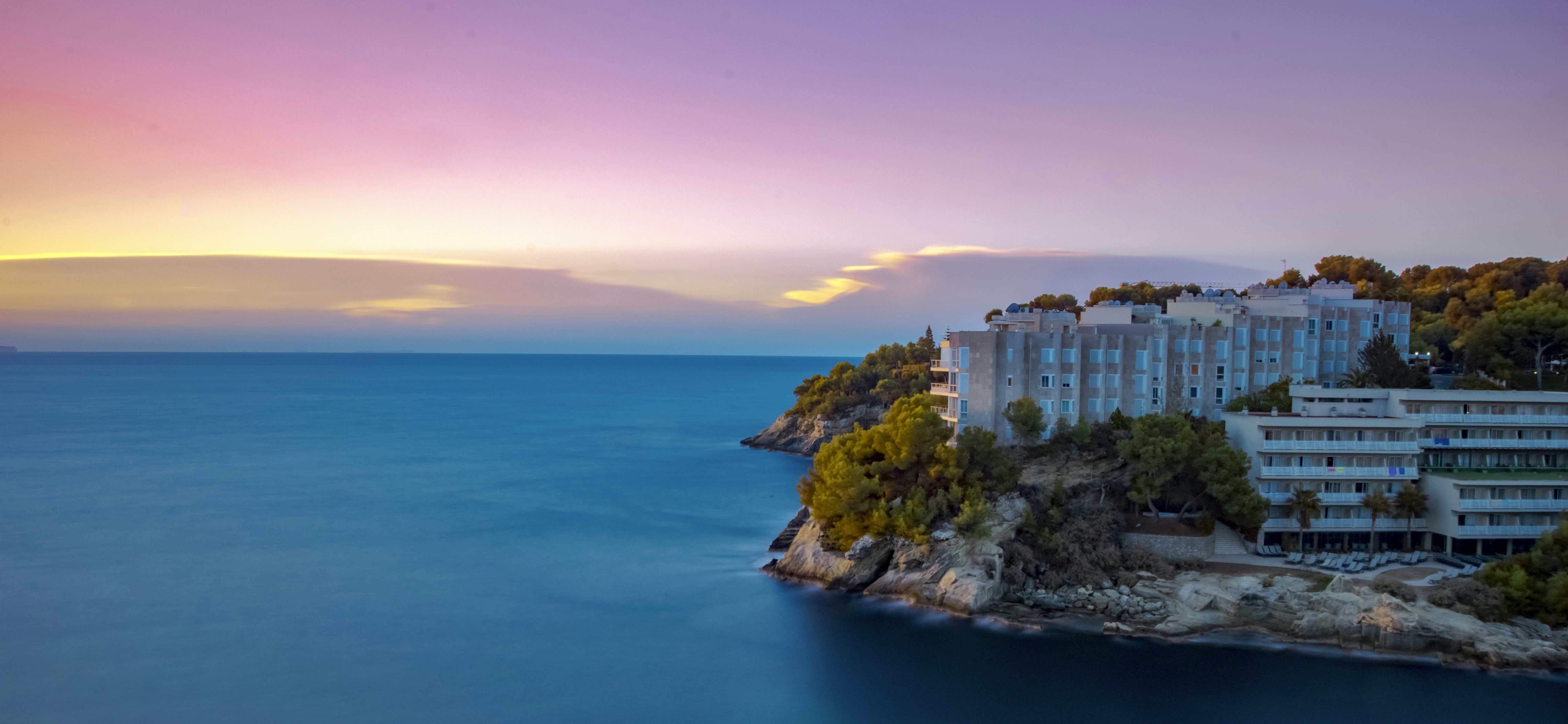 Coastal hotel perched on a rocky cliff overlooking the calm, expansive ocean at sunset.