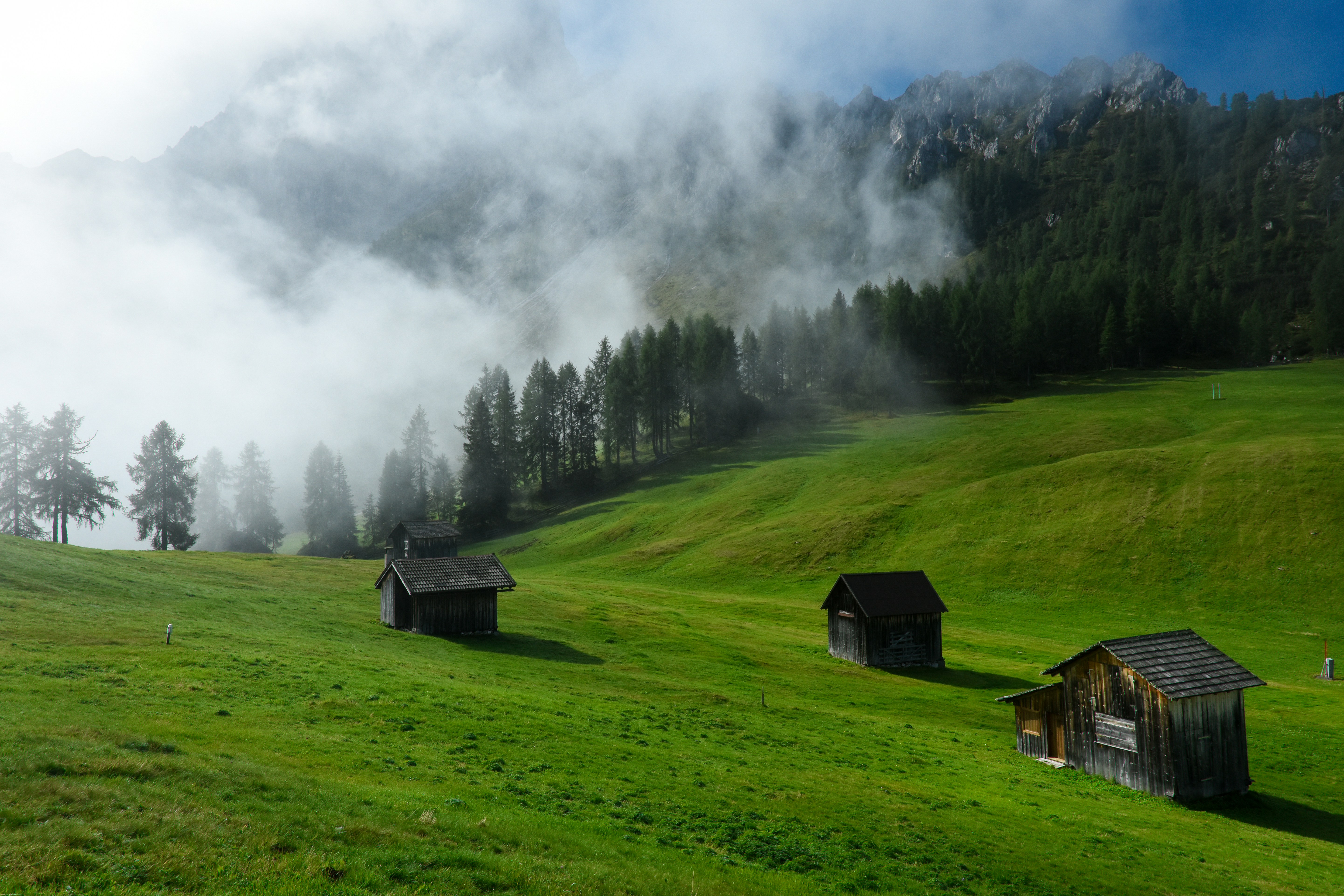Rustic cabins dot a lush green hillside beneath a veil of misty clouds and distant forested mountains.