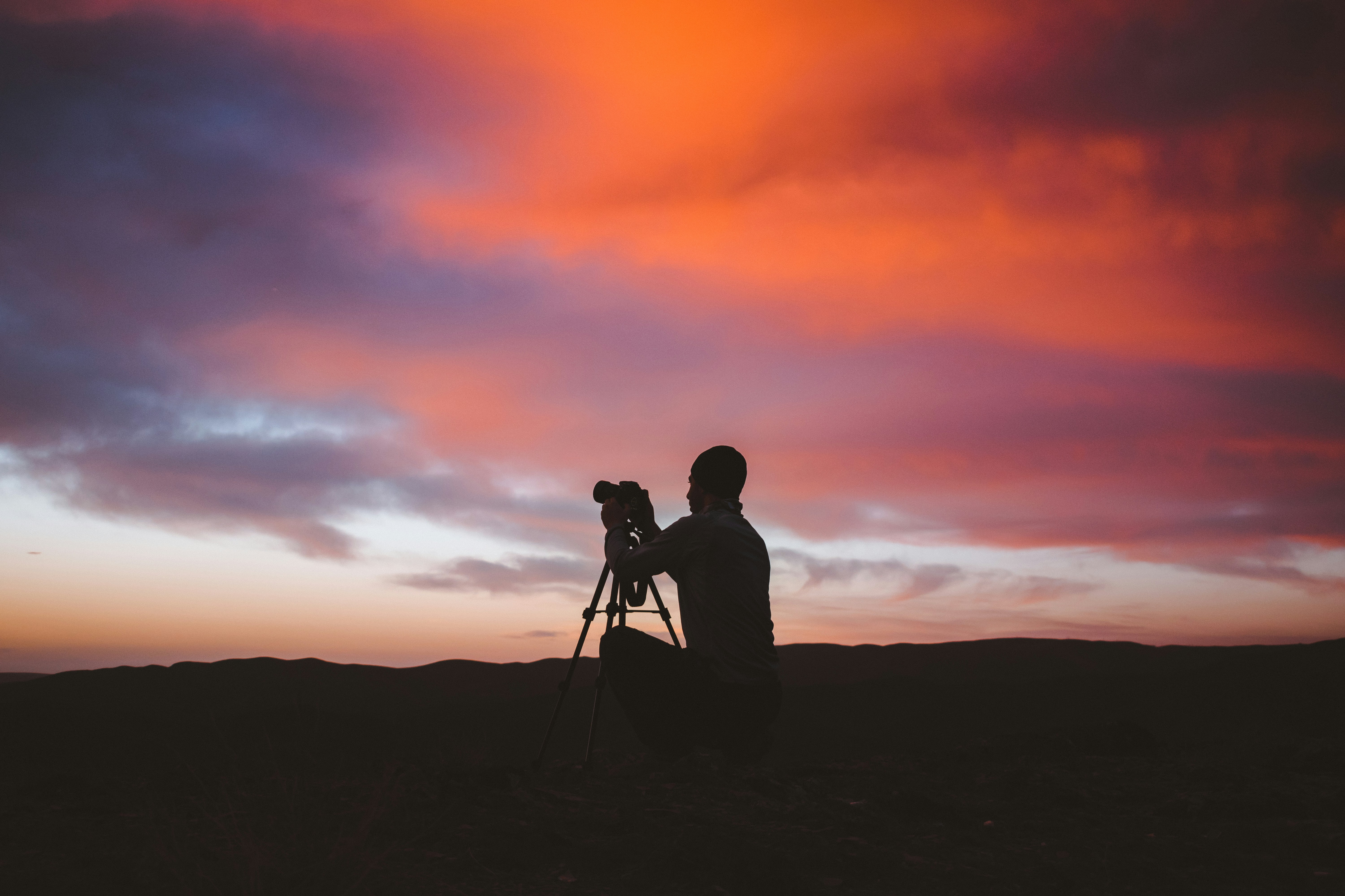 Silhouette of a photographer capturing a vibrant sunset, framed by dramatic clouds and distant hills.