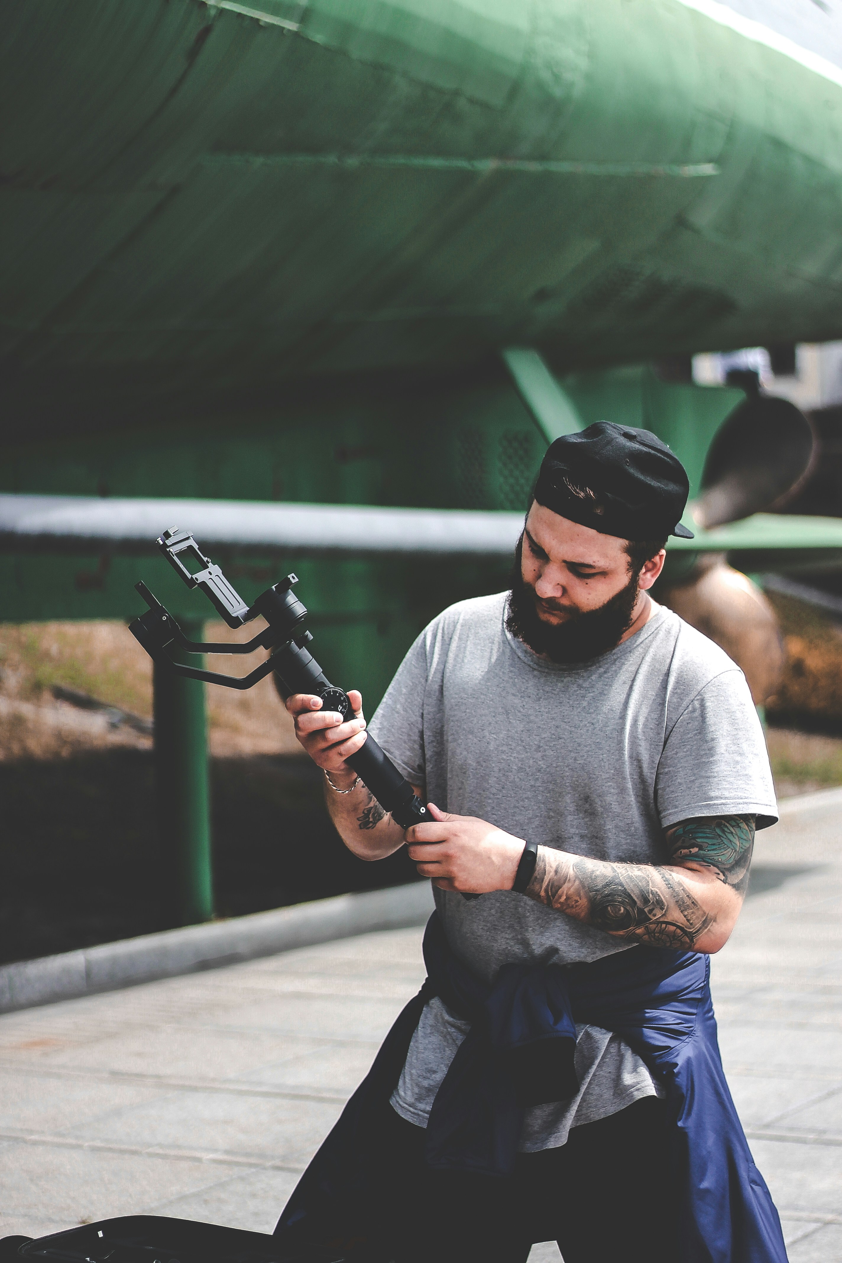 A man adjusts a camera stabilizer, focused on his task in an outdoor setting with a vintage aircraft in the background.