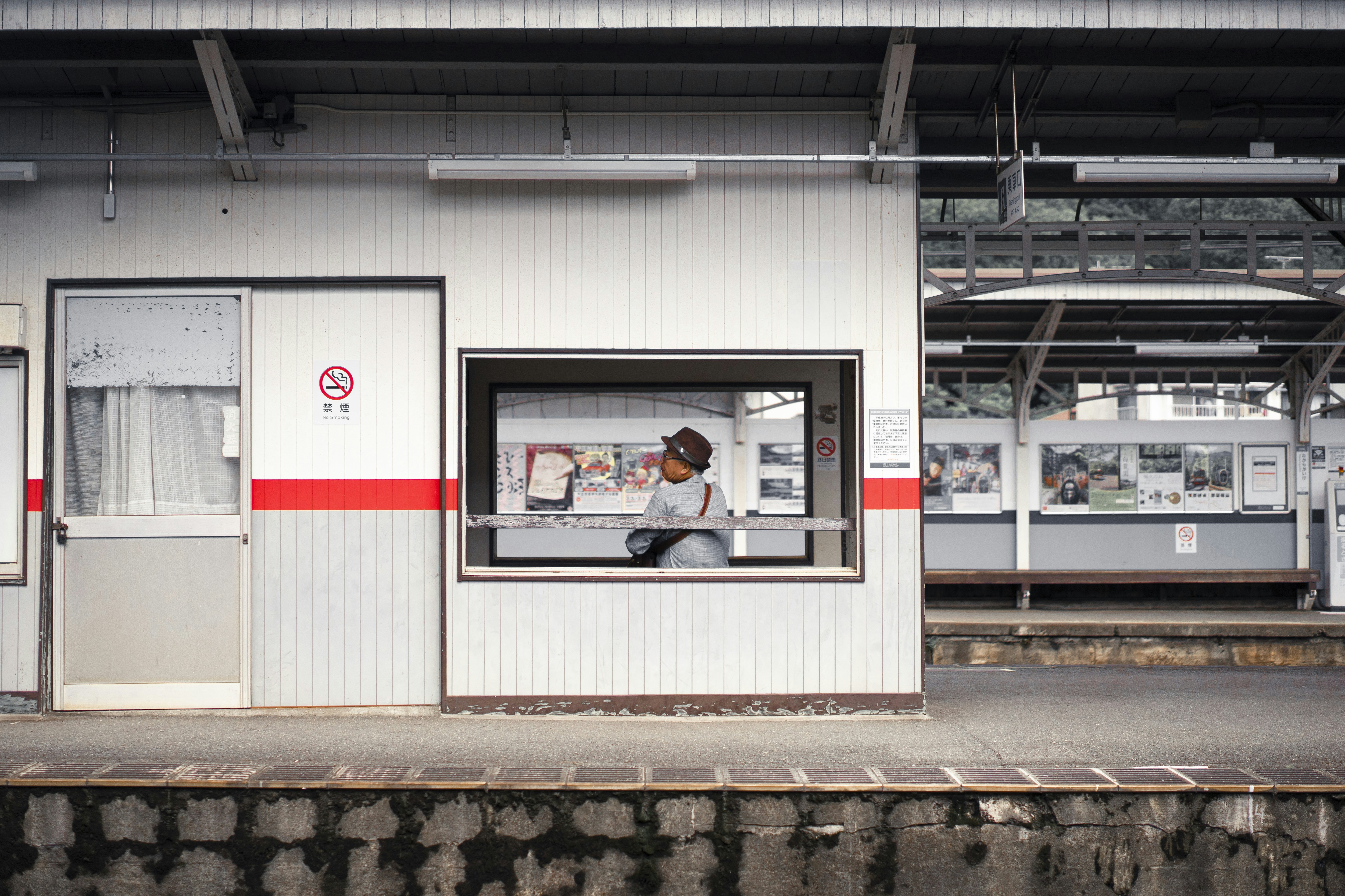 A lone figure sits in a train station window, framed by minimalist architecture and subtle color accents. The scene evokes a sense of waiting and contemplation.