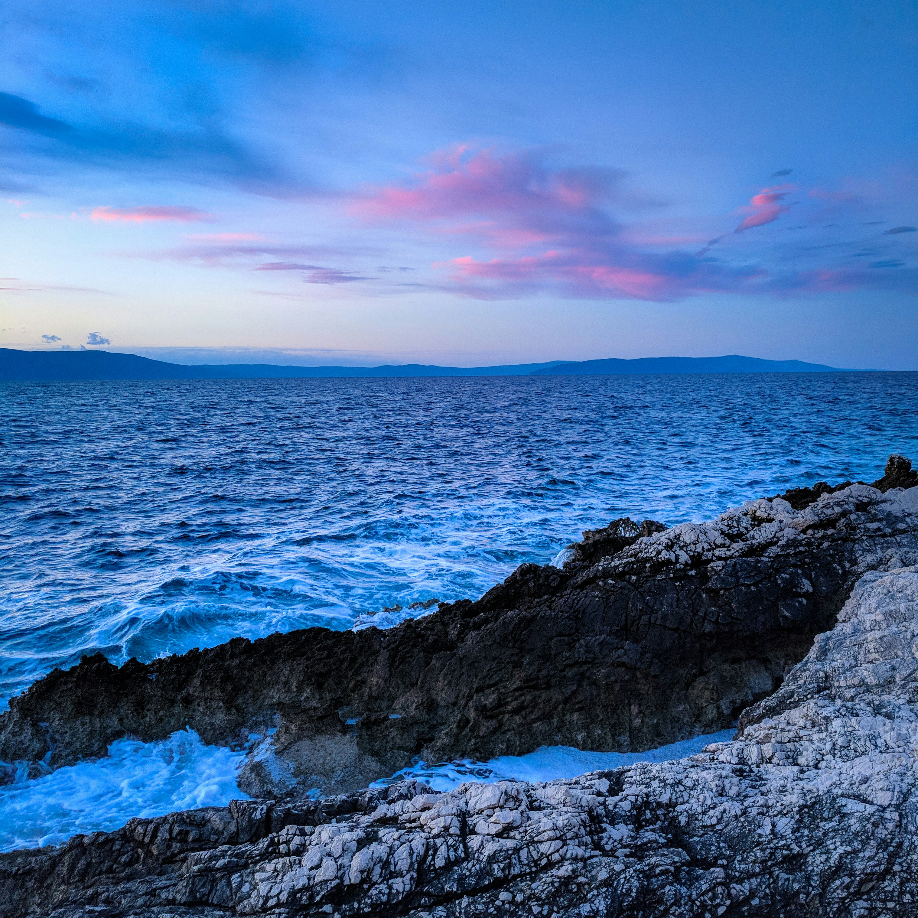 Vibrant pink clouds hover over a rugged coastline as waves crash against the rocks at dusk.
