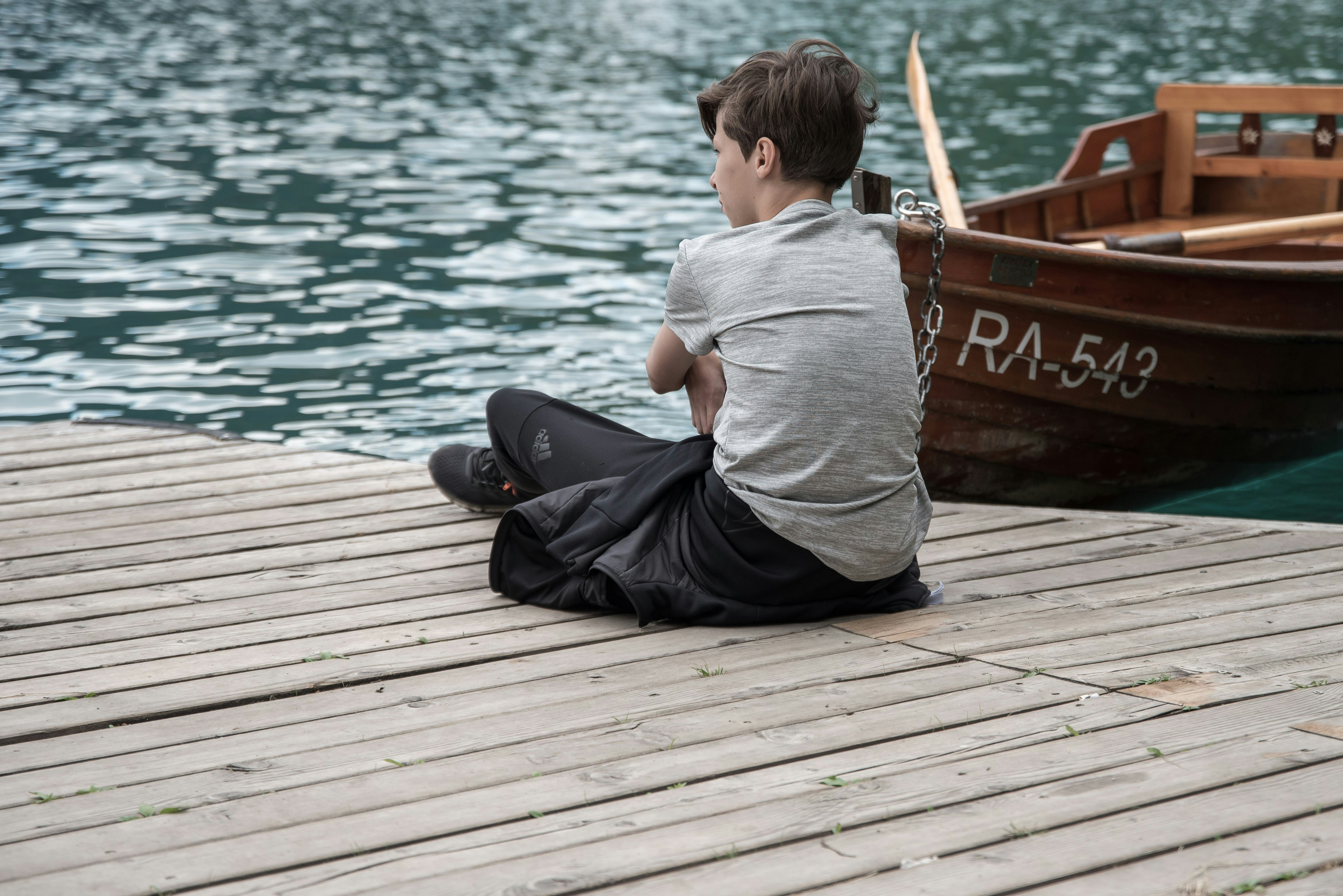Young boy sits pensively on a wooden dock beside a rustic boat on a tranquil lake.