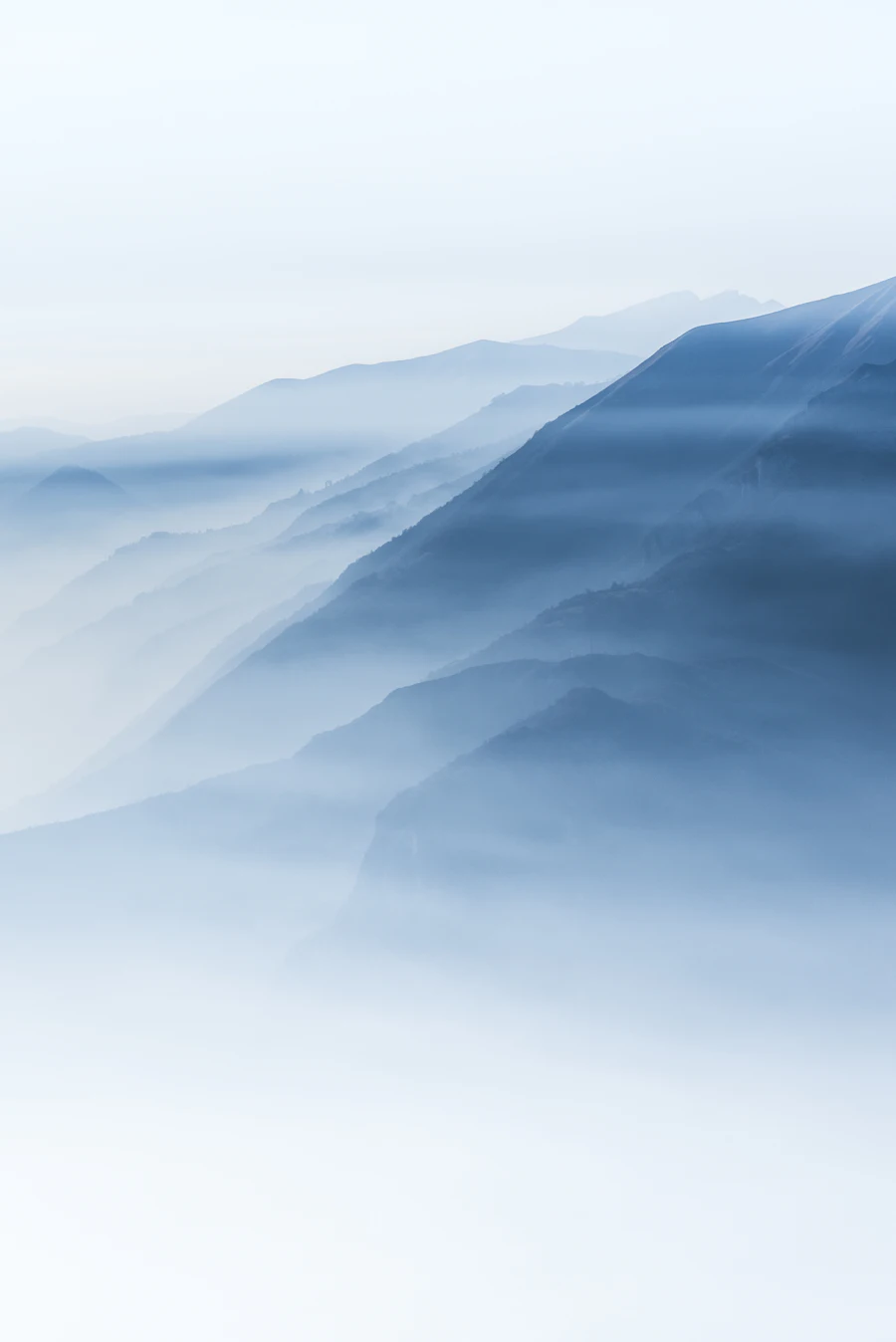 Layered mountain ridges fading into pale blue haze, with a large dark slope cutting diagonally through the right side. Soft fog fills the valleys and bright sky washes the top of the frame.