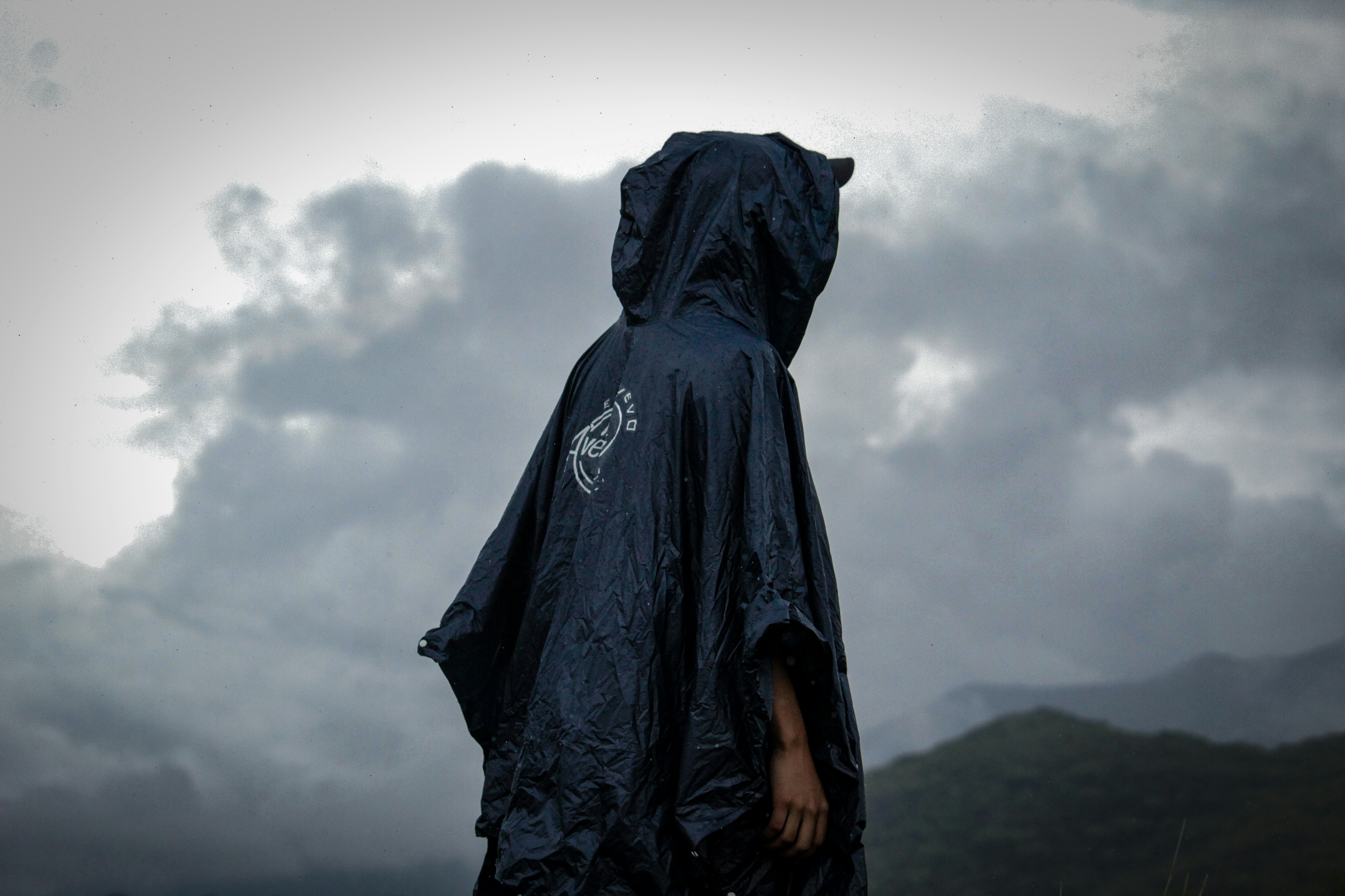Figure in a dark poncho stands against a backdrop of dramatic, overcast clouds.