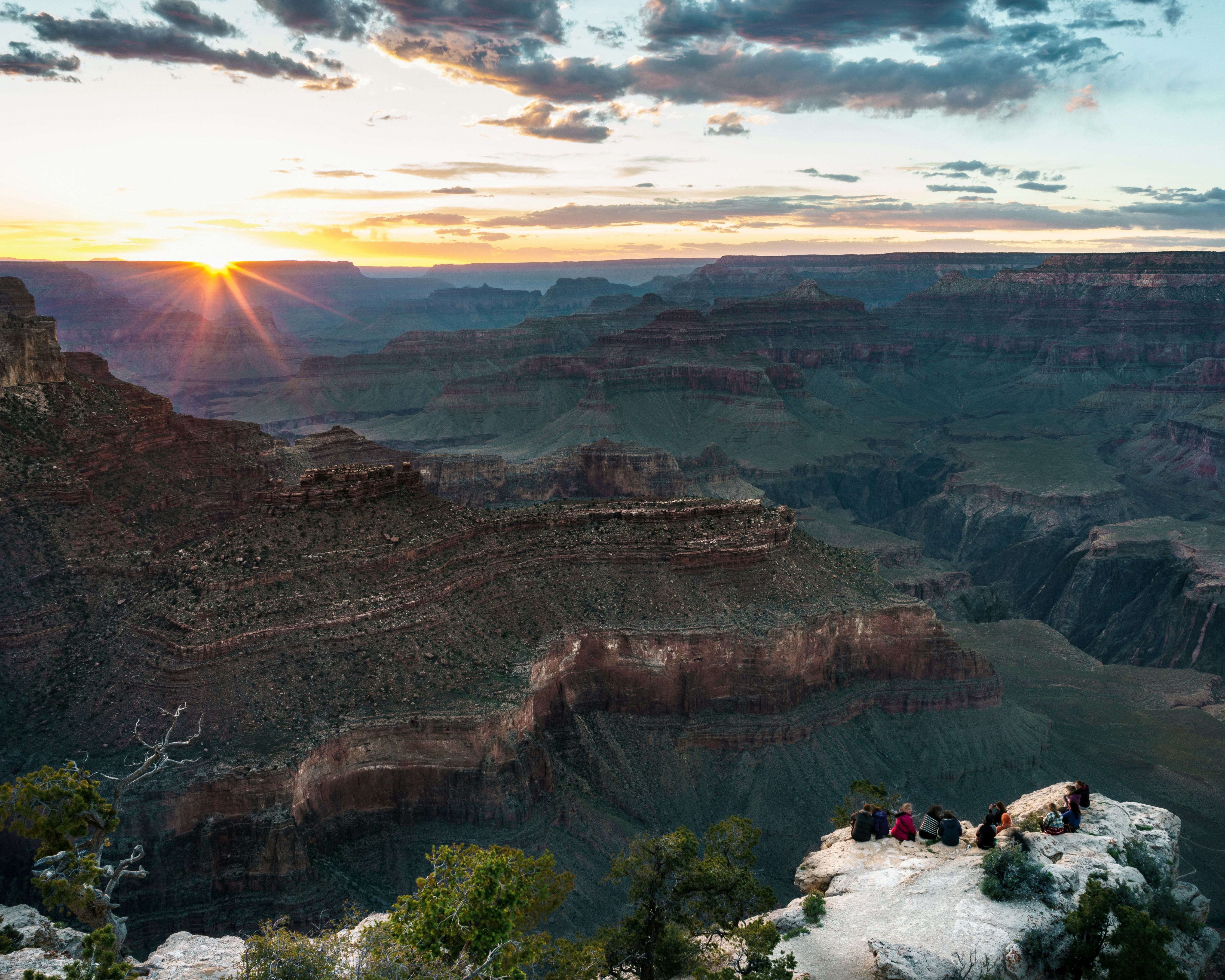 Silhouetted figures sitting on a rocky ledge as the sun sets over the expansive Grand Canyon, highlighting the rugged terrain and vibrant sky.