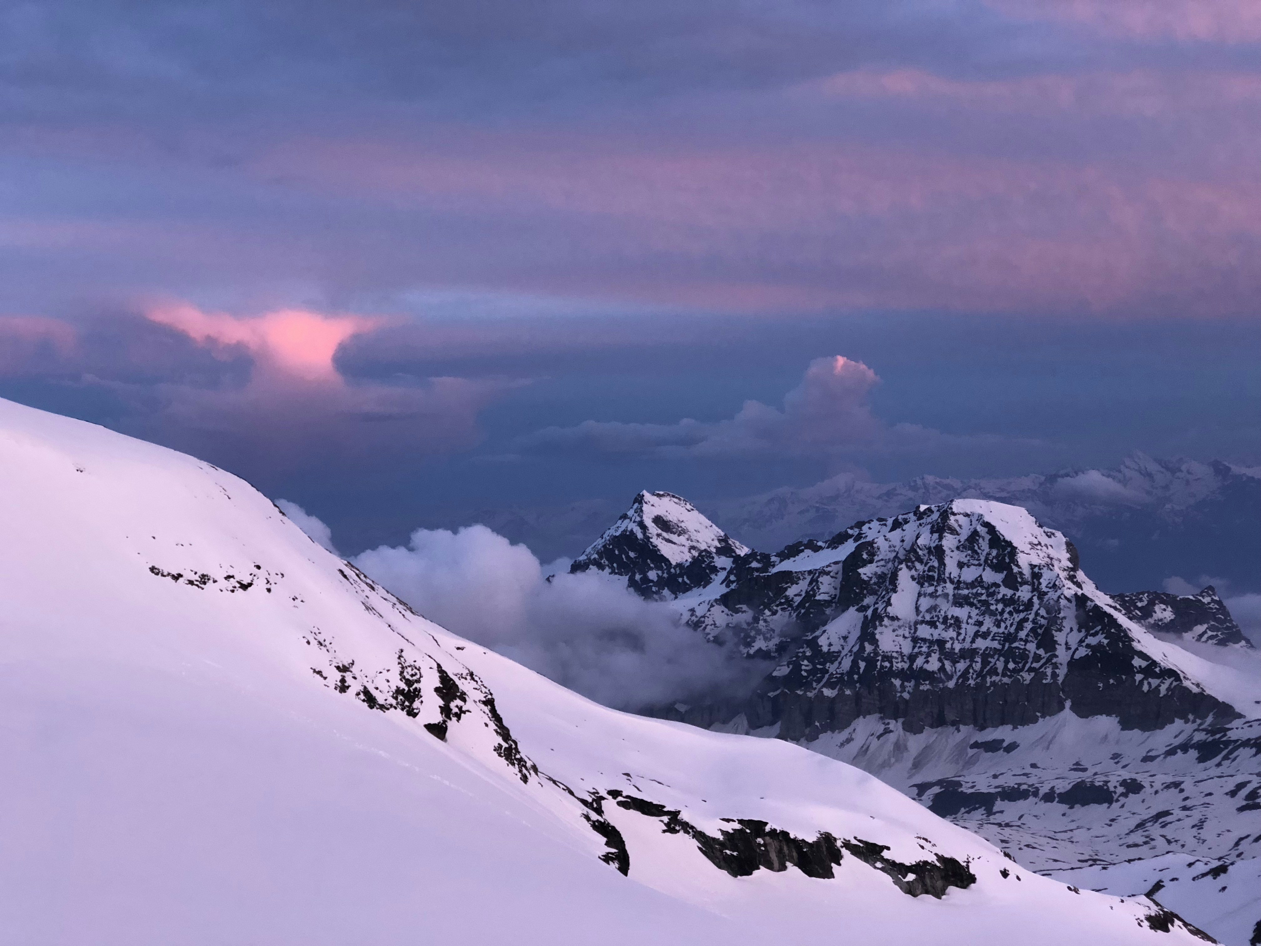 Majestic snow-covered peaks rise against a pastel sky, hinting at the transition from day to night. Clouds weave through the valleys, adding depth to the serene landscape.