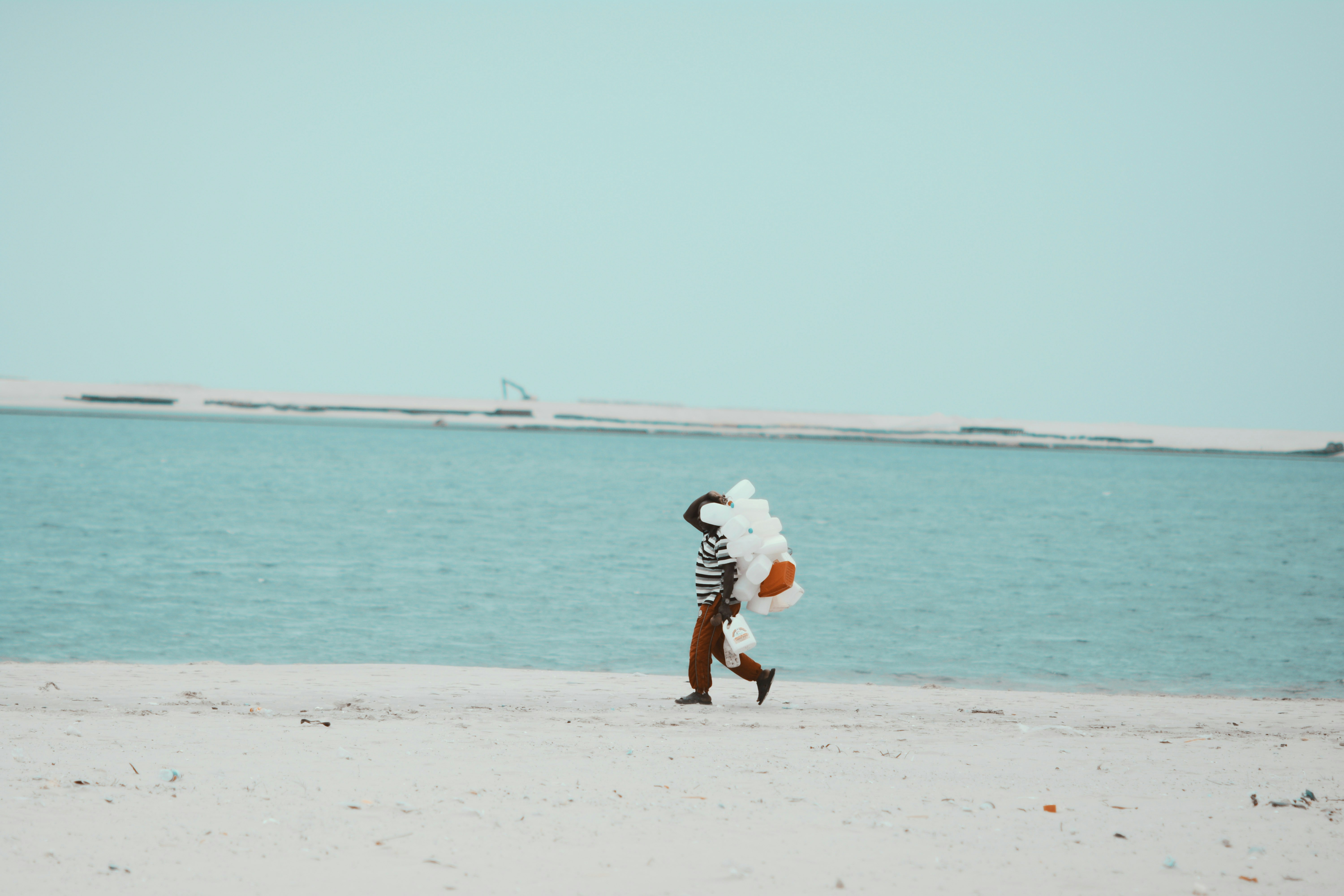 Person walking along a sandy beach carrying a keg with a turquoise sea in the background.