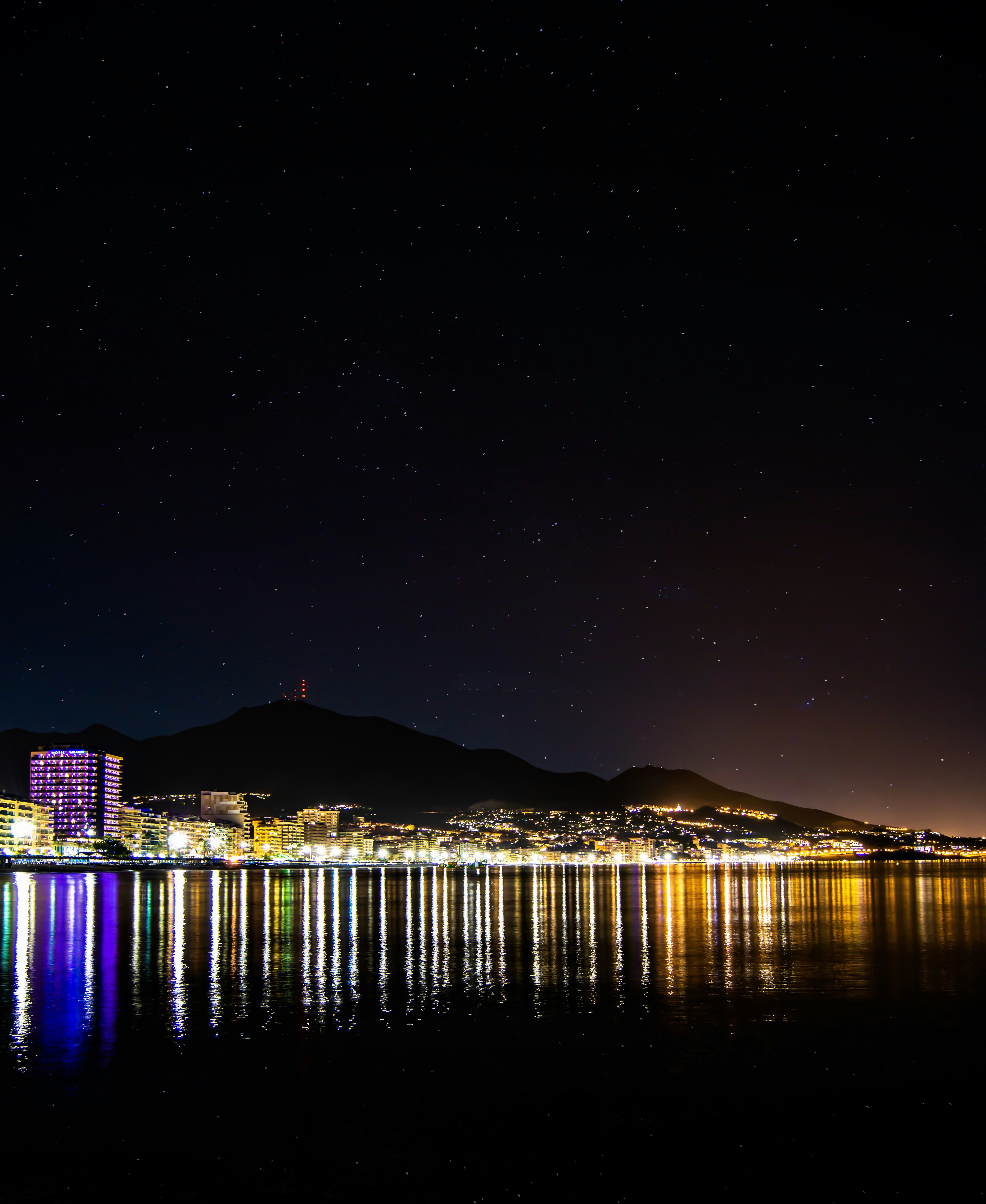 Vibrant city lights reflecting on calm waters under a starry night sky, with a silhouette of mountains in the background.