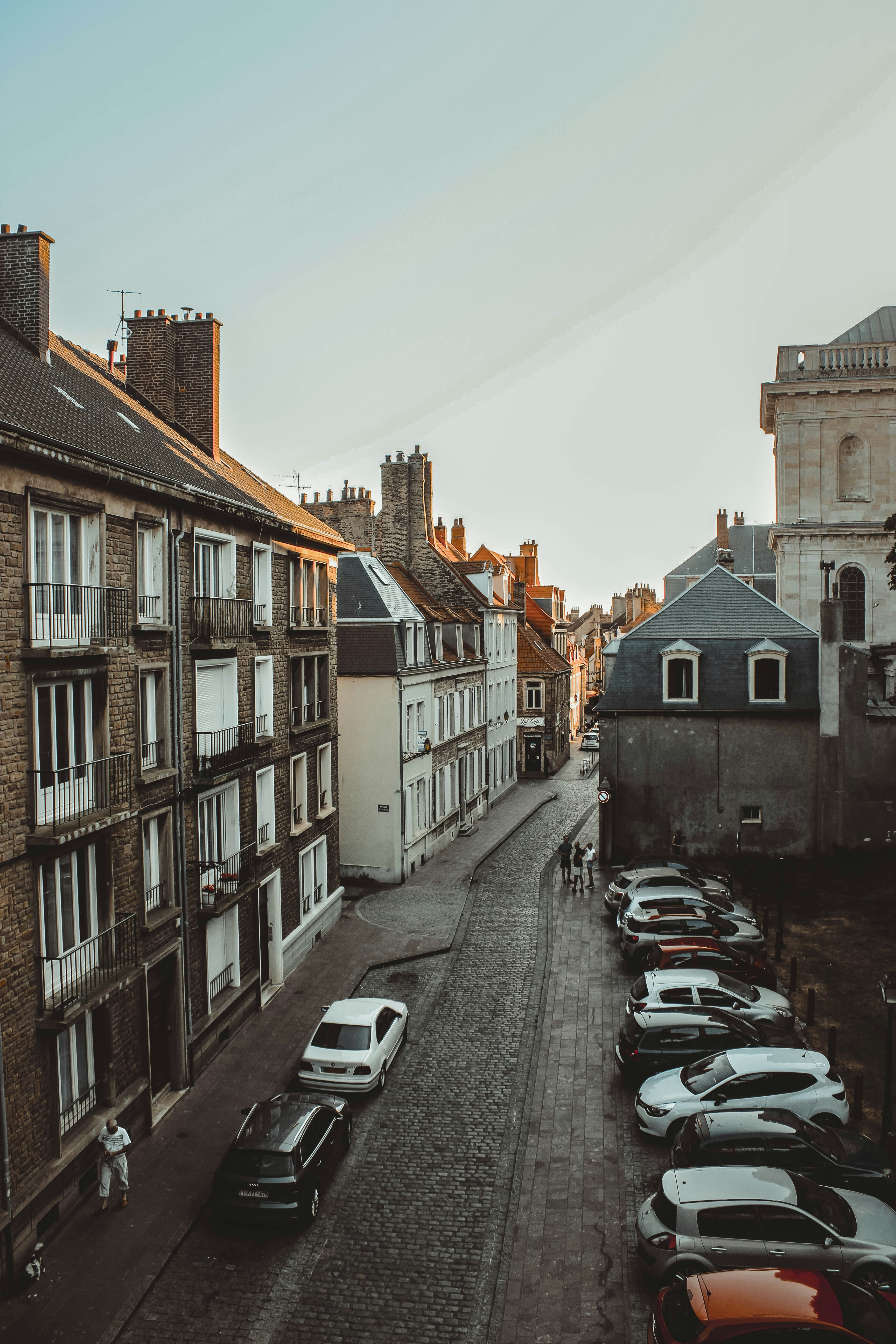 Charming cobblestone street lined with historic buildings and parked cars, bathed in warm evening light. A few pedestrians stroll along the lane.
