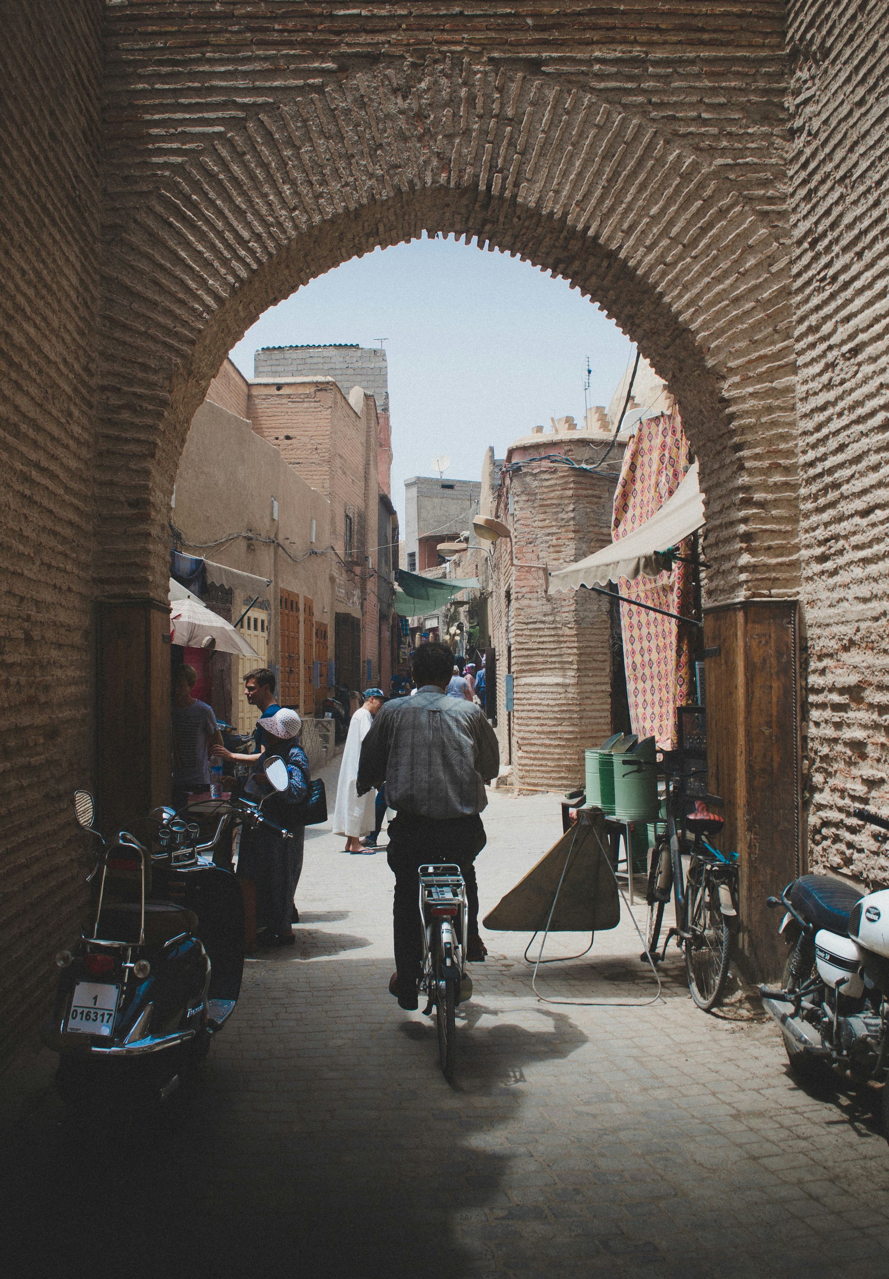 A cyclist navigates a bustling market street framed by an ancient arch, with local vendors and bicycles lining the path.