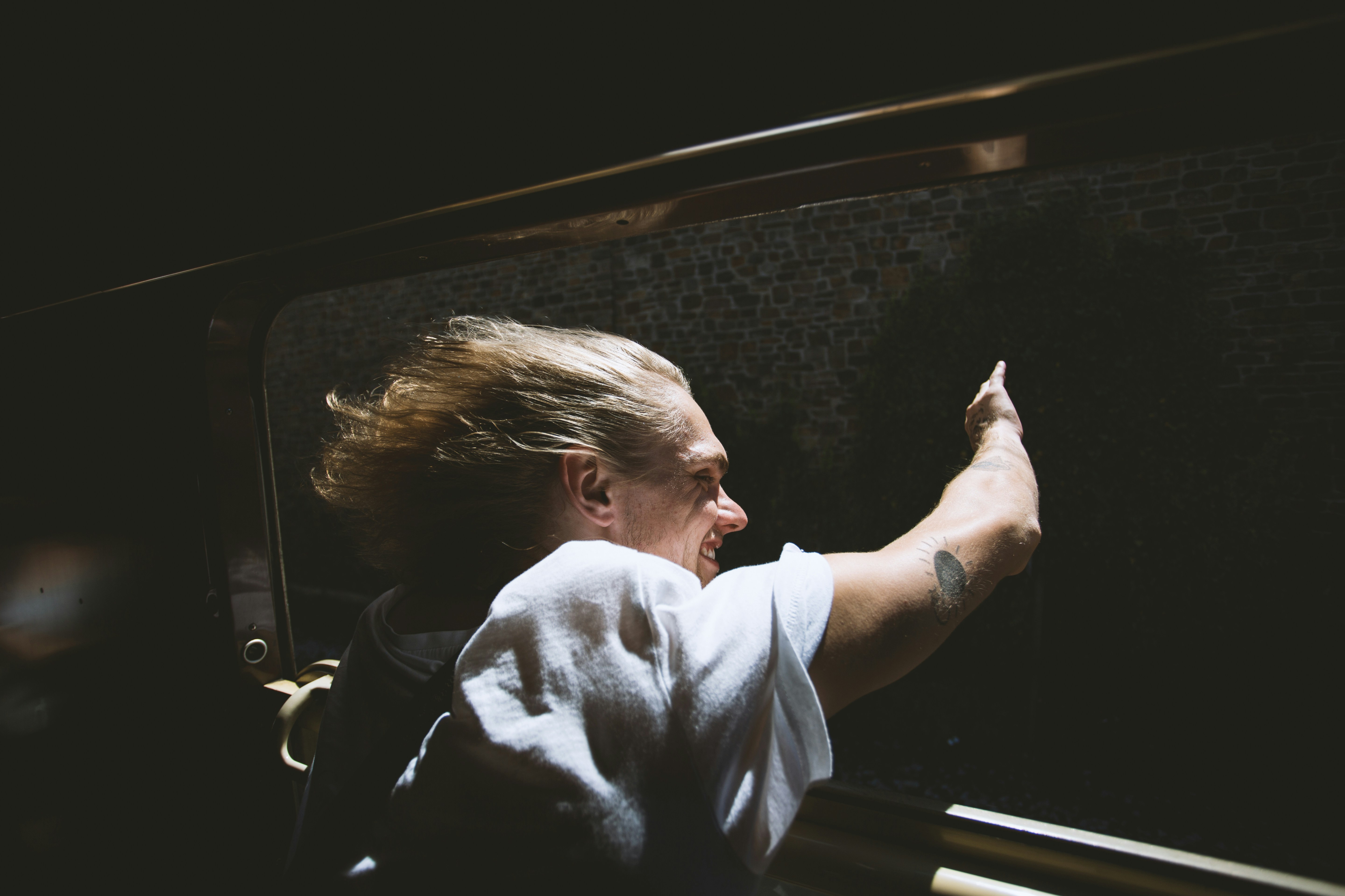 Individual joyfully extending their arm out of a vehicle window, hair flowing in the wind. Natural light highlights the carefree spirit of the scene.