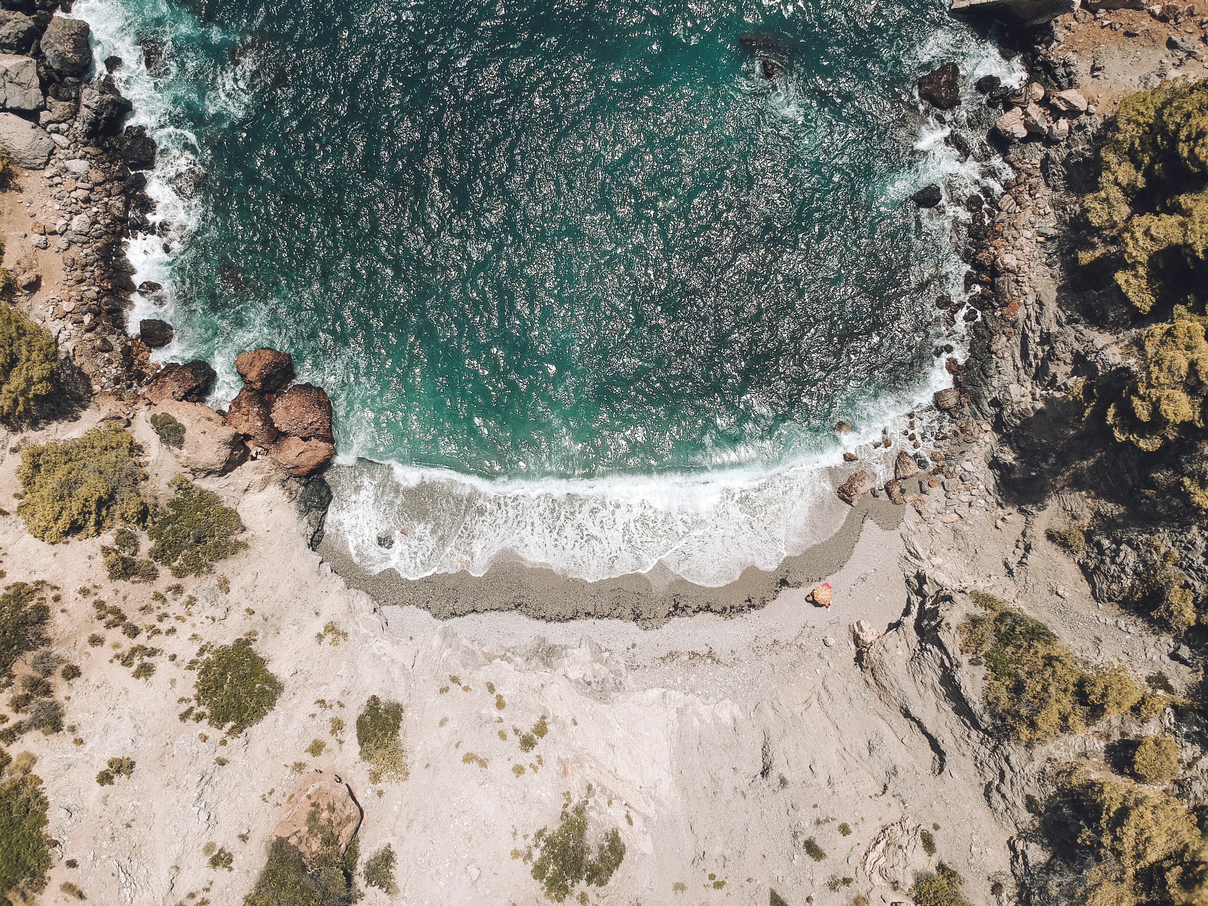 Aerial view of a secluded cove with turquoise waters meeting a sandy shoreline, surrounded by rocky outcrops and sparse vegetation.