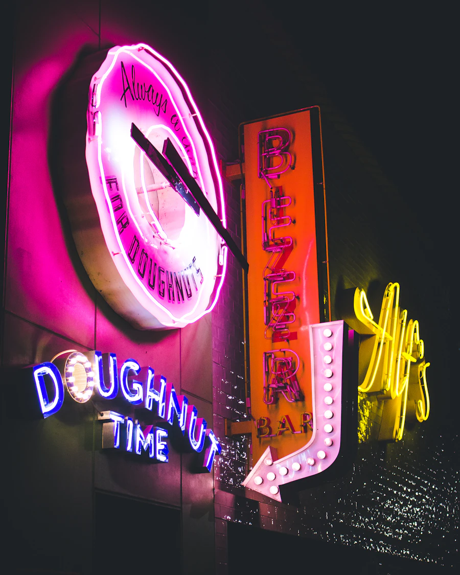 Bright neon signs for a doughnut shop at night, including a pink circular sign, a vertical orange beer sign, and blue “Doughnut Time” lettering on a dark storefront.