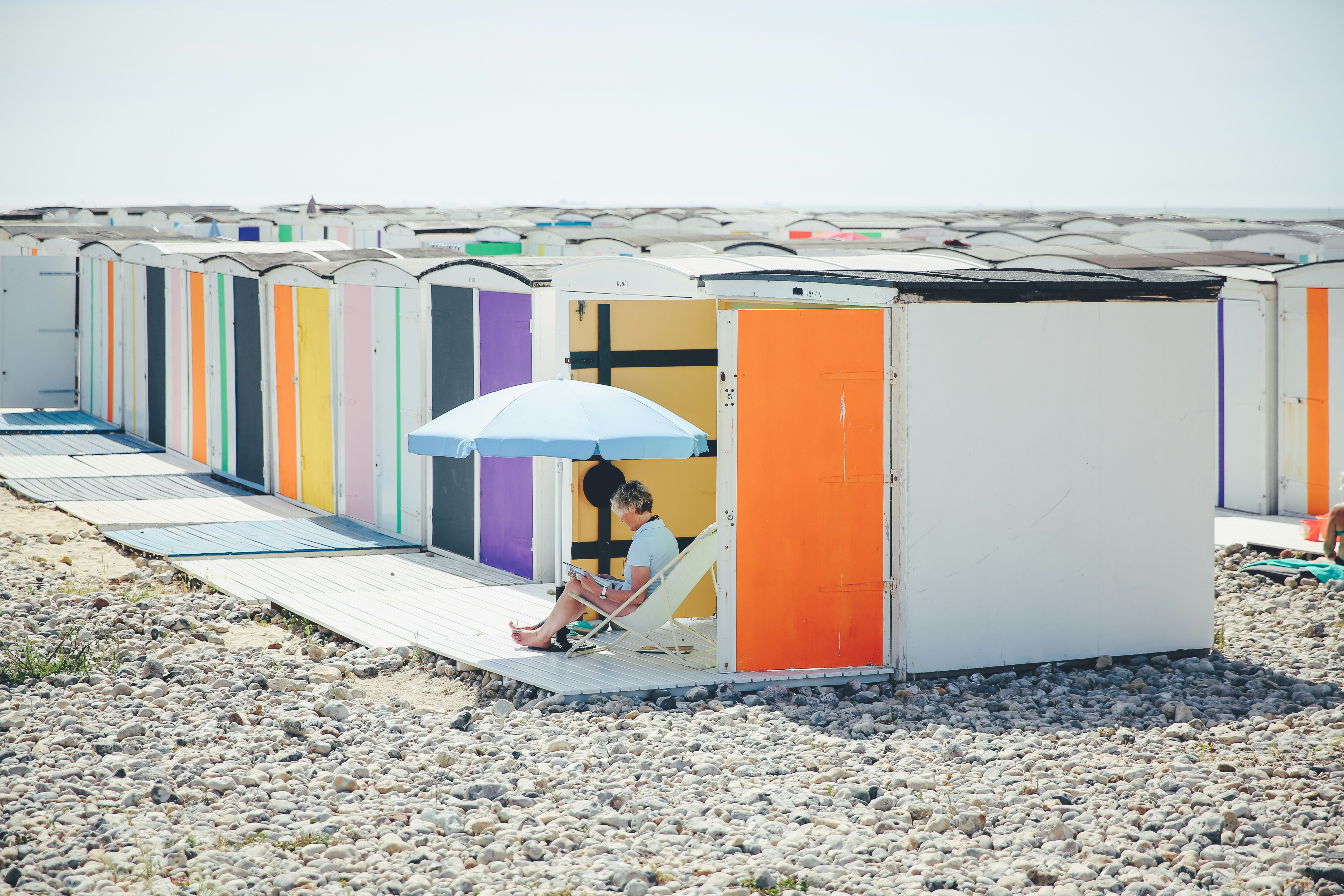 Colorful beach huts with a person sitting under a blue umbrella on a pebble shoreline.
