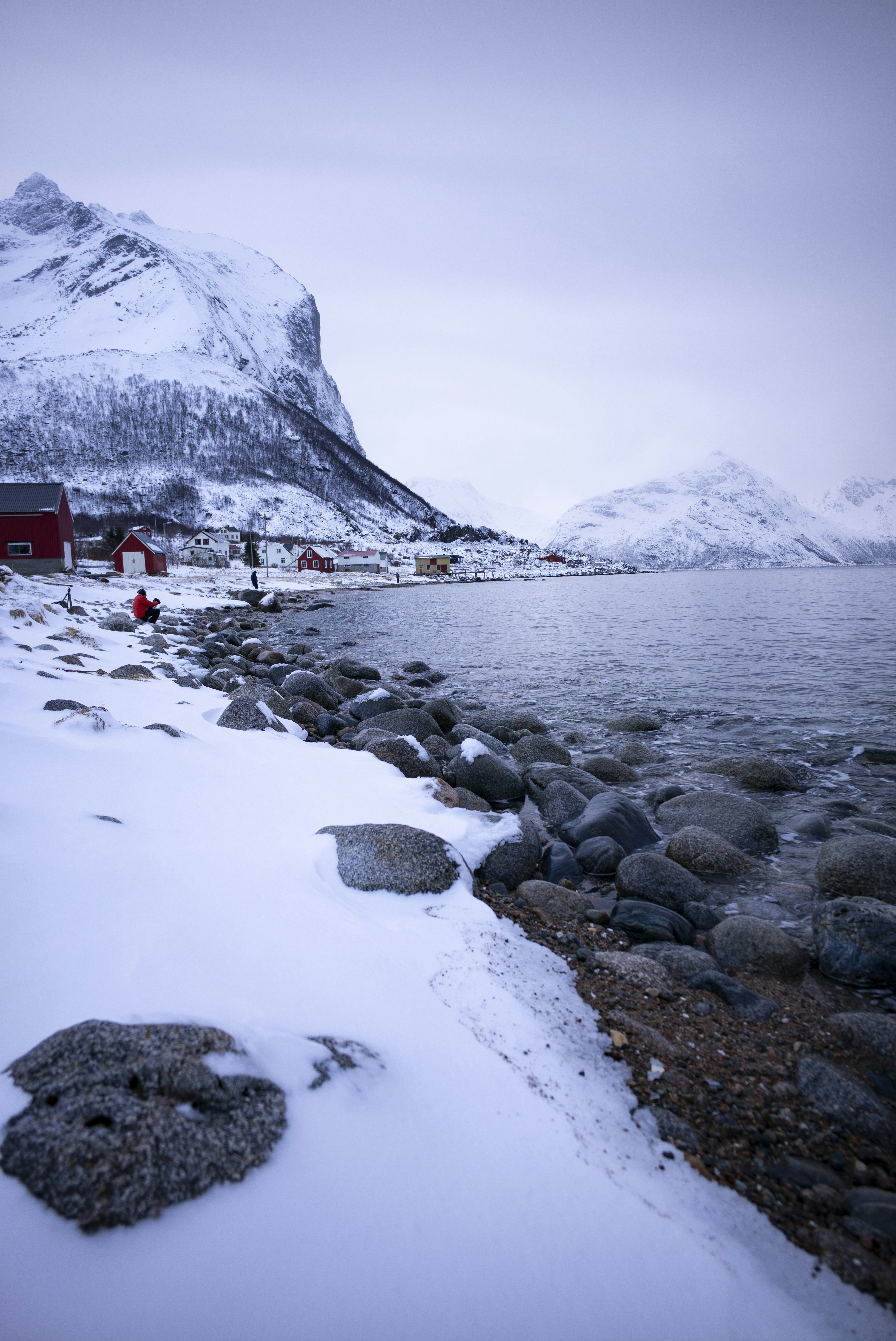 A tranquil winter scene featuring a snowy shoreline with scattered rocks and a distant village nestled against towering mountains.