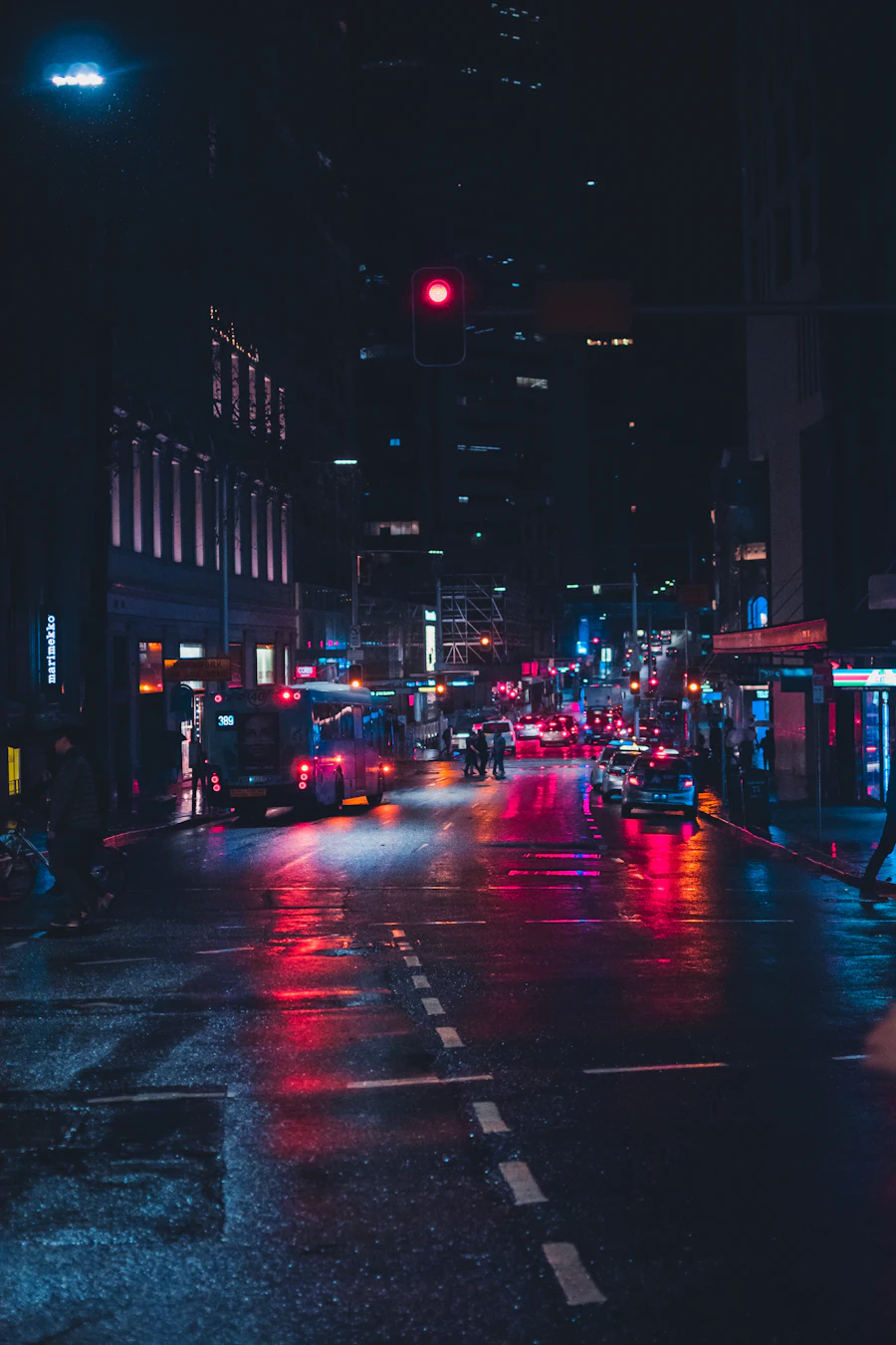 Wet city street at night glowing with red and blue neon reflections, with a red traffic light above the road and cars moving through the scene.