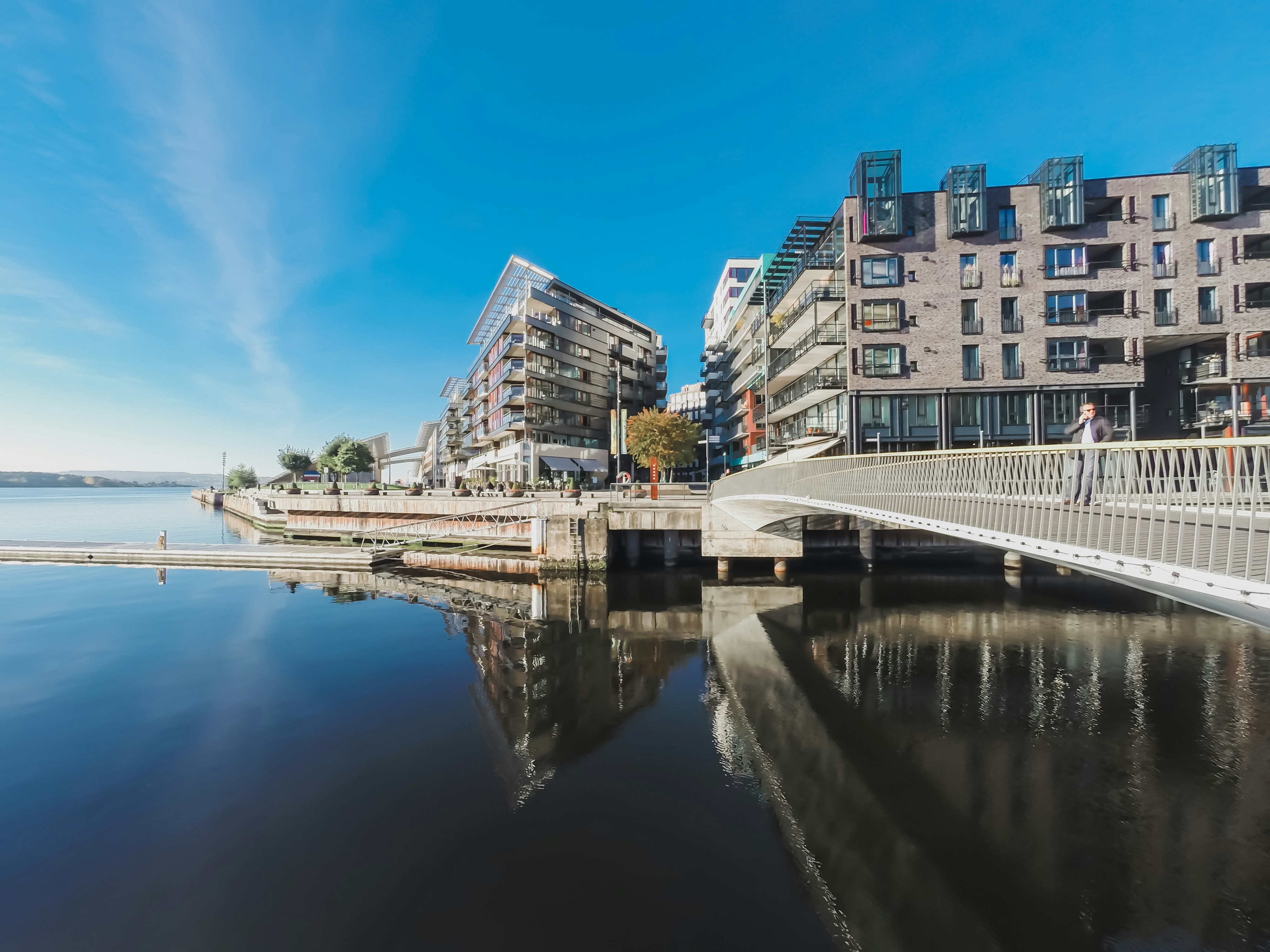 Modern bridge spanning a calm river with reflections of city buildings under a clear blue sky.