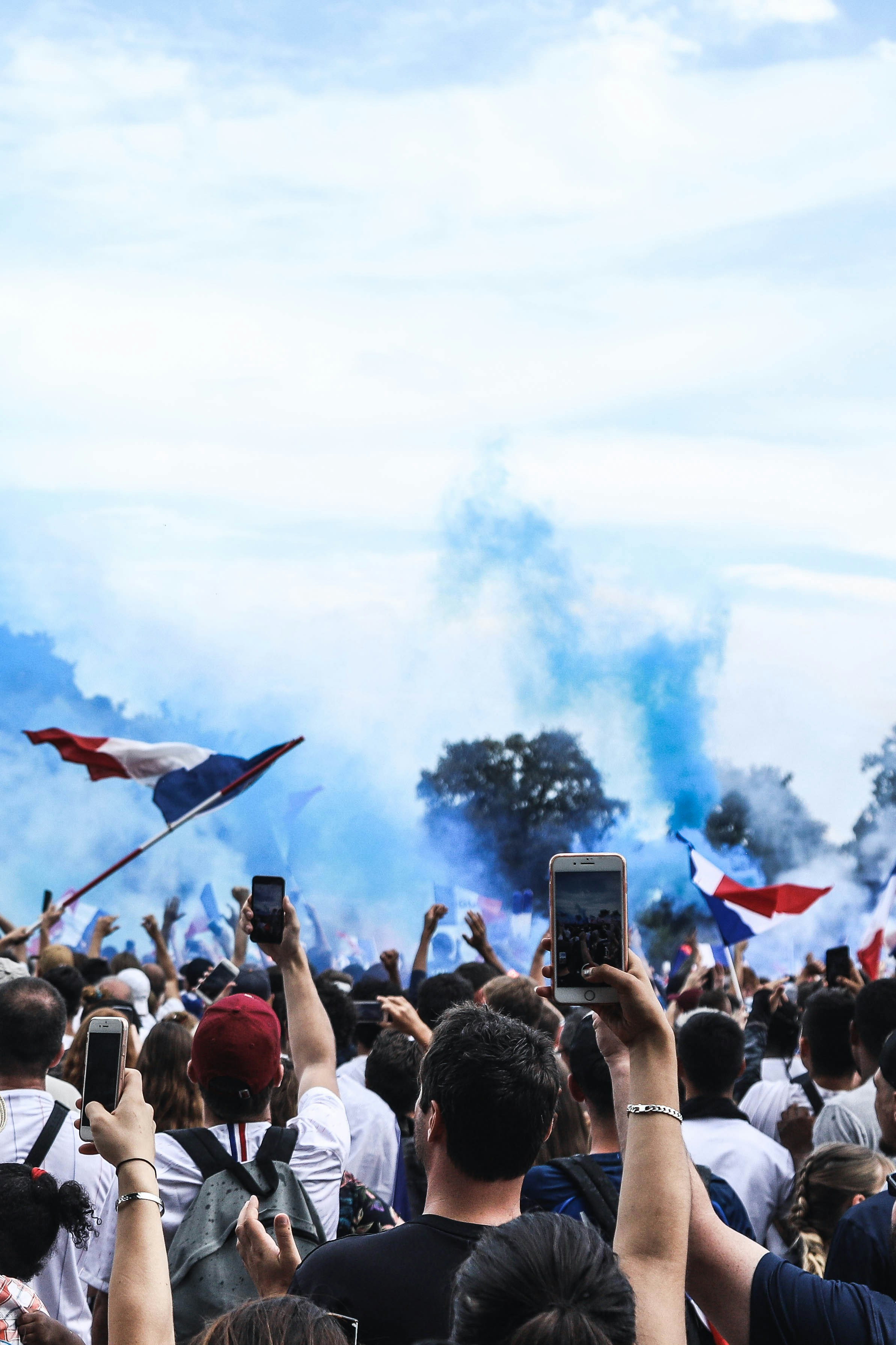 Crowd of enthusiastic supporters waving flags and capturing the moment with smartphones, surrounded by colorful smoke in the air.