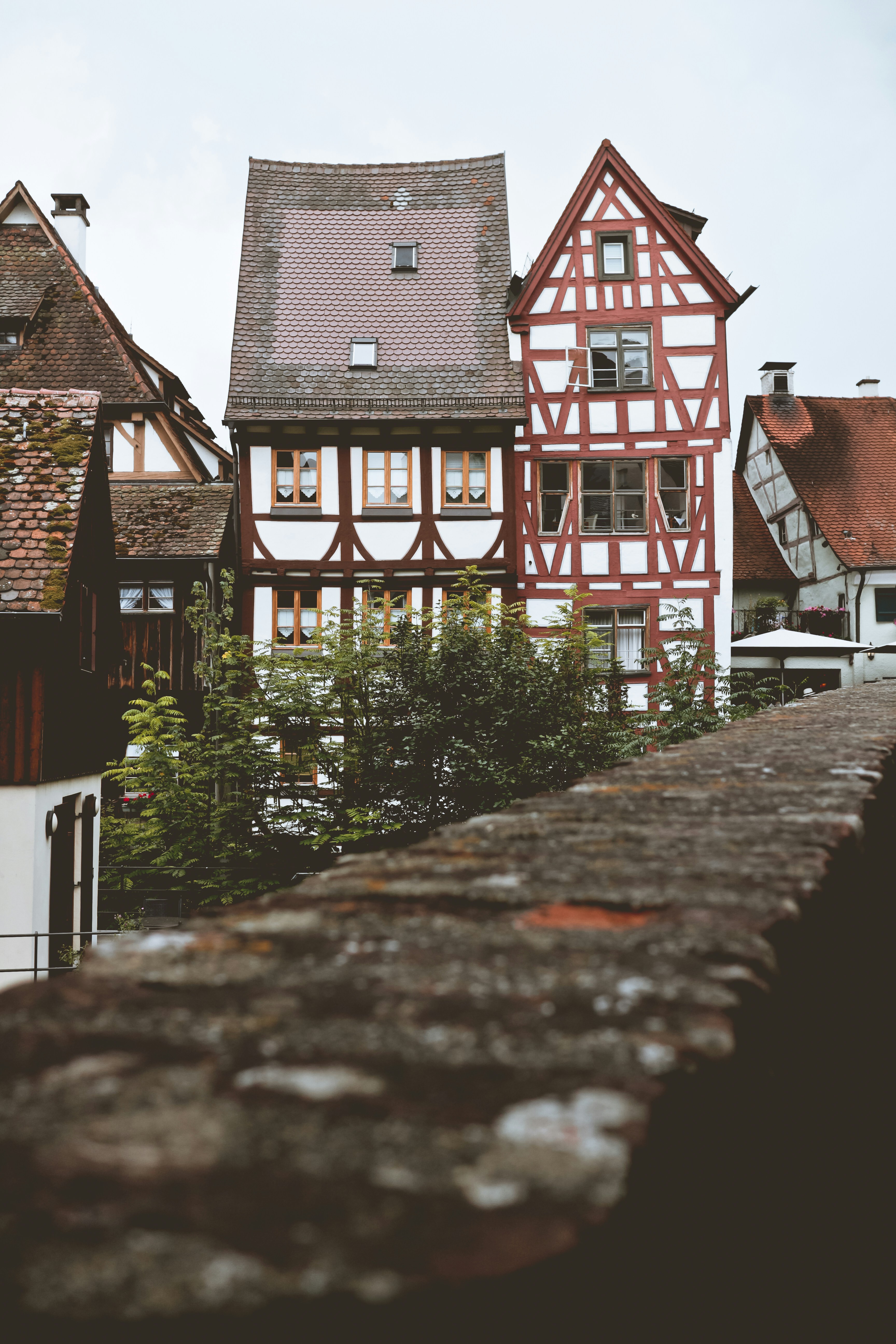 Row of half-timbered houses with red framing rises behind a mossy stone wall under a pale sky.