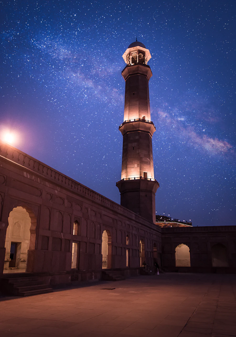 Tall illuminated minaret rising above a courtyard at night, with a bright star-filled sky and the Milky Way arcing behind it. Warm lights glow from the building’s arches below.