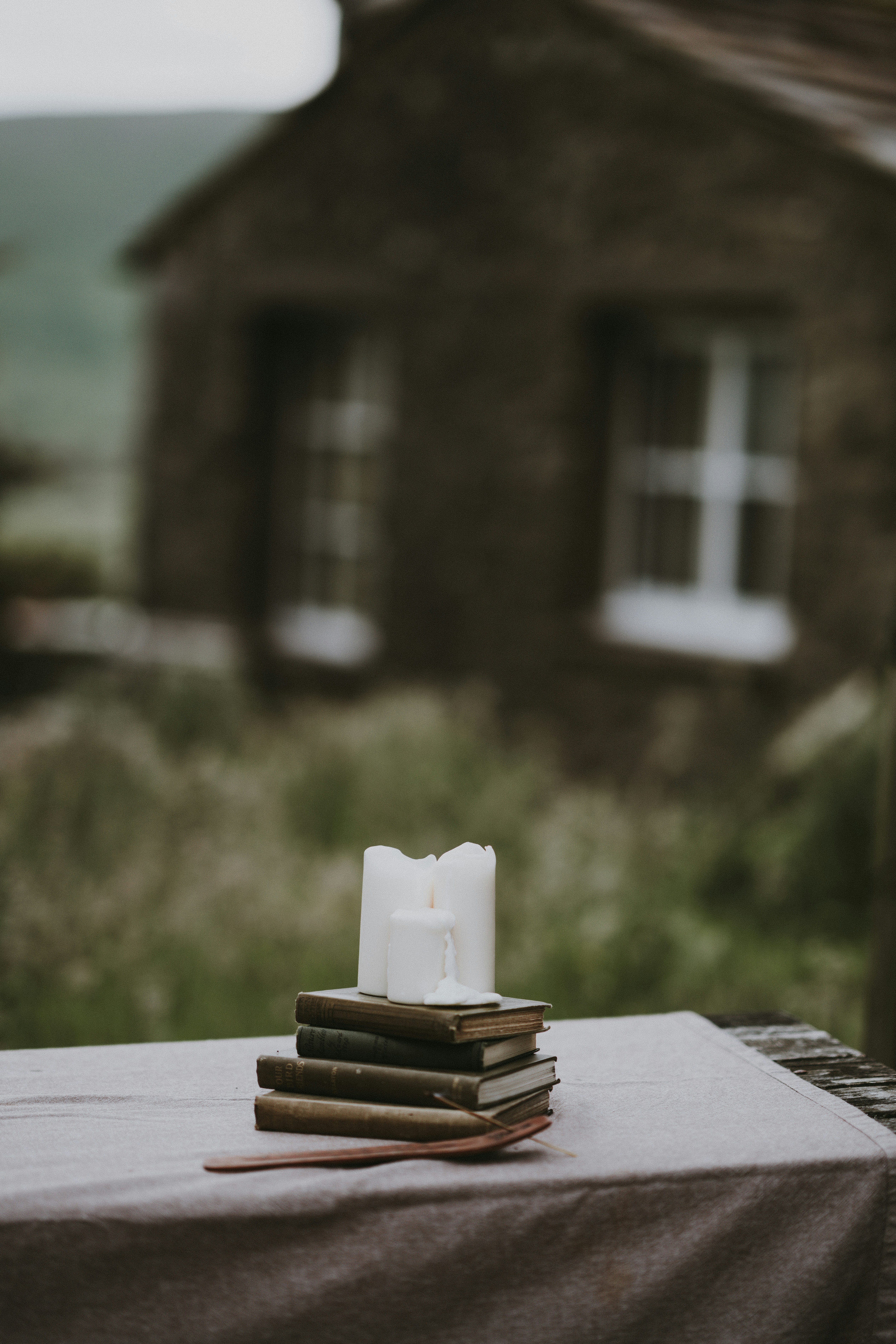 Stack of vintage books topped with lit candles on a table, set against a blurred stone house backdrop. 