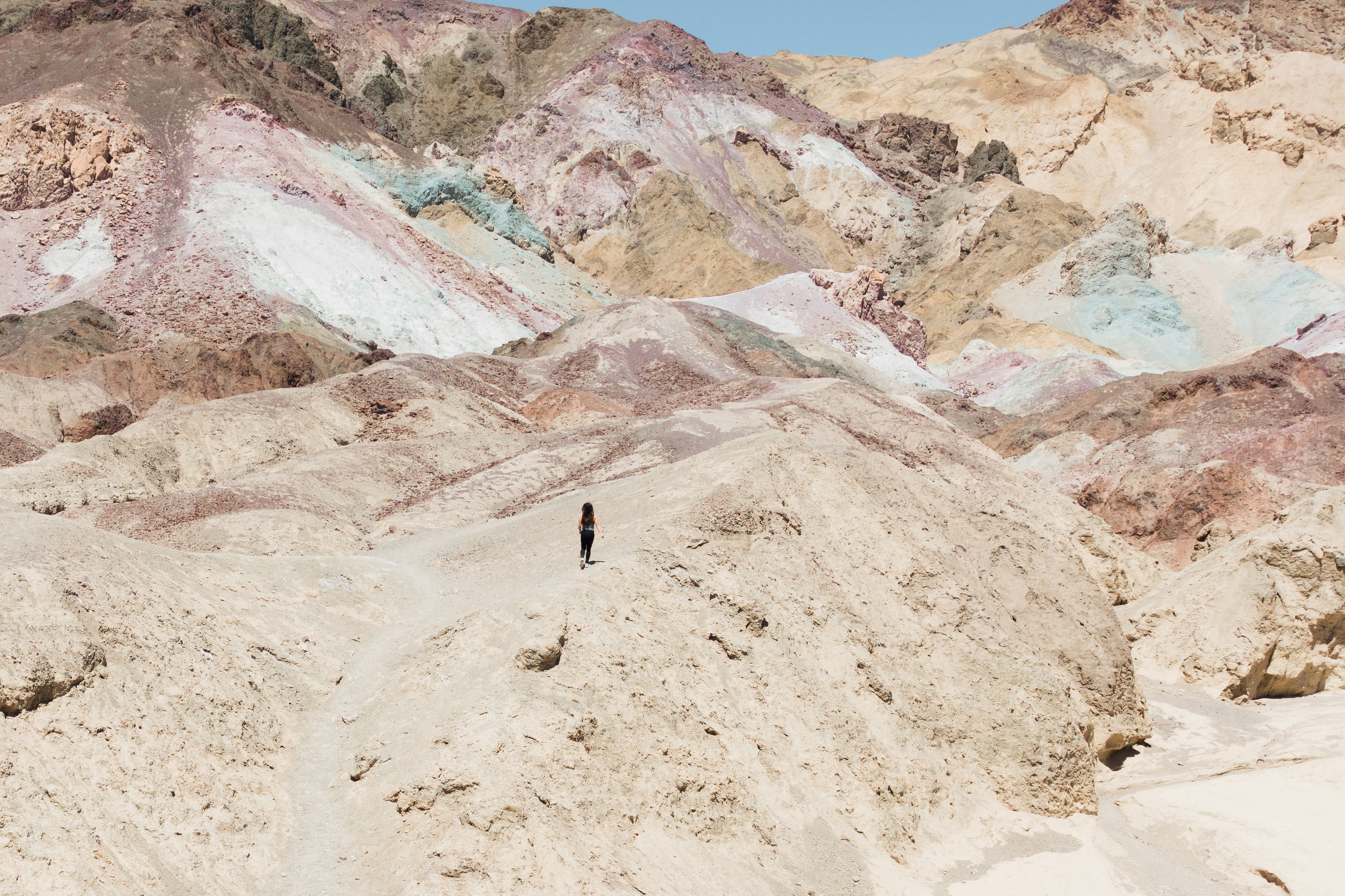 Person walking on a path amidst colorful, layered hills under a clear blue sky.