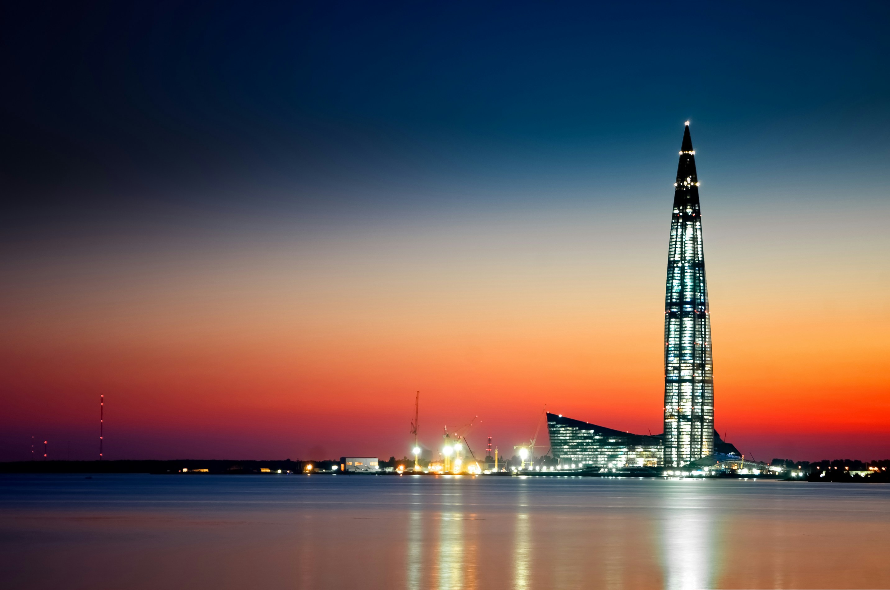 Tall skyscraper illuminated against a vivid sunset gradient over calm waters.